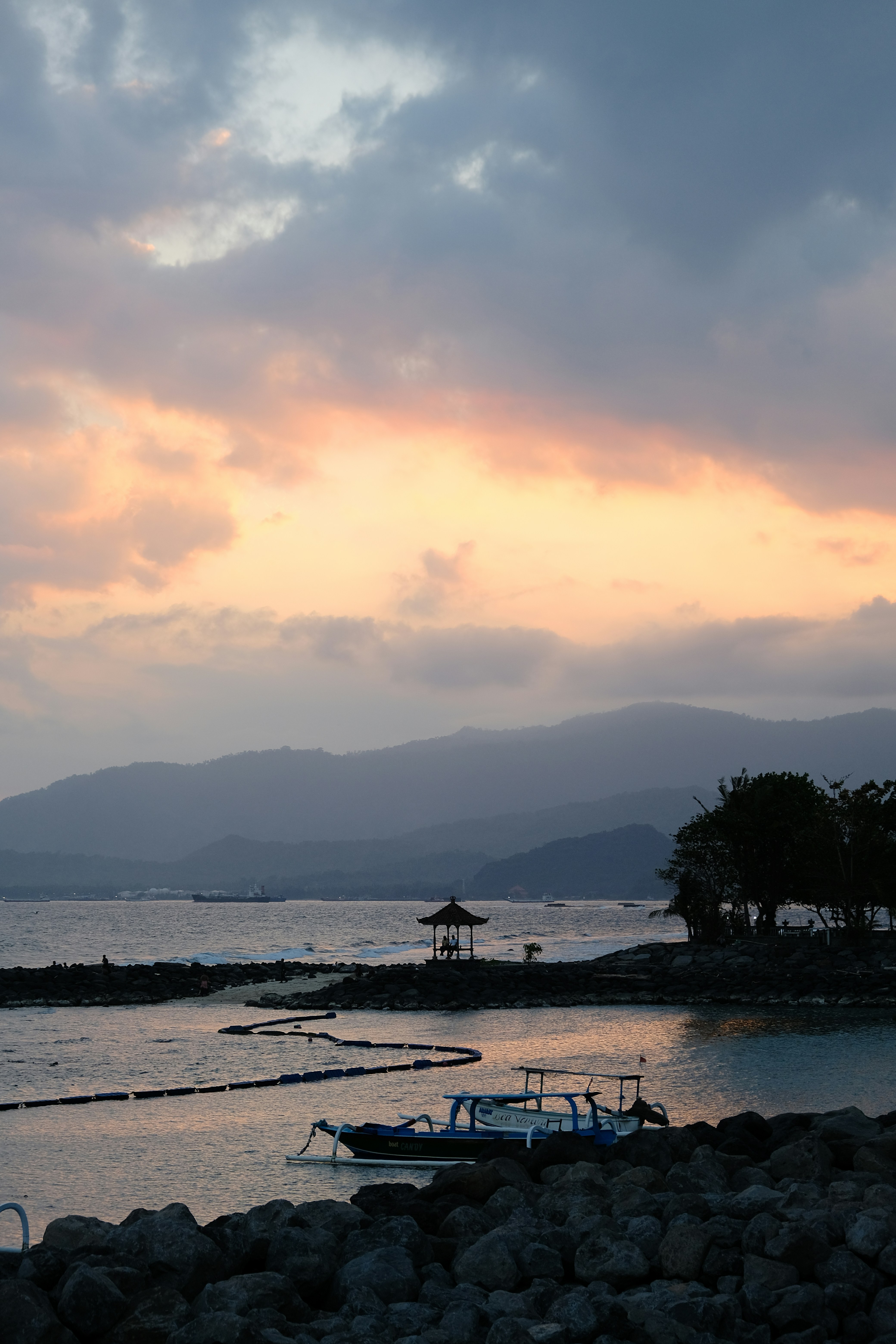 Silhouetted gazebo and fishing boats reflect the tranquility of a coastal sunset, framed by distant mountains and a serene shoreline.