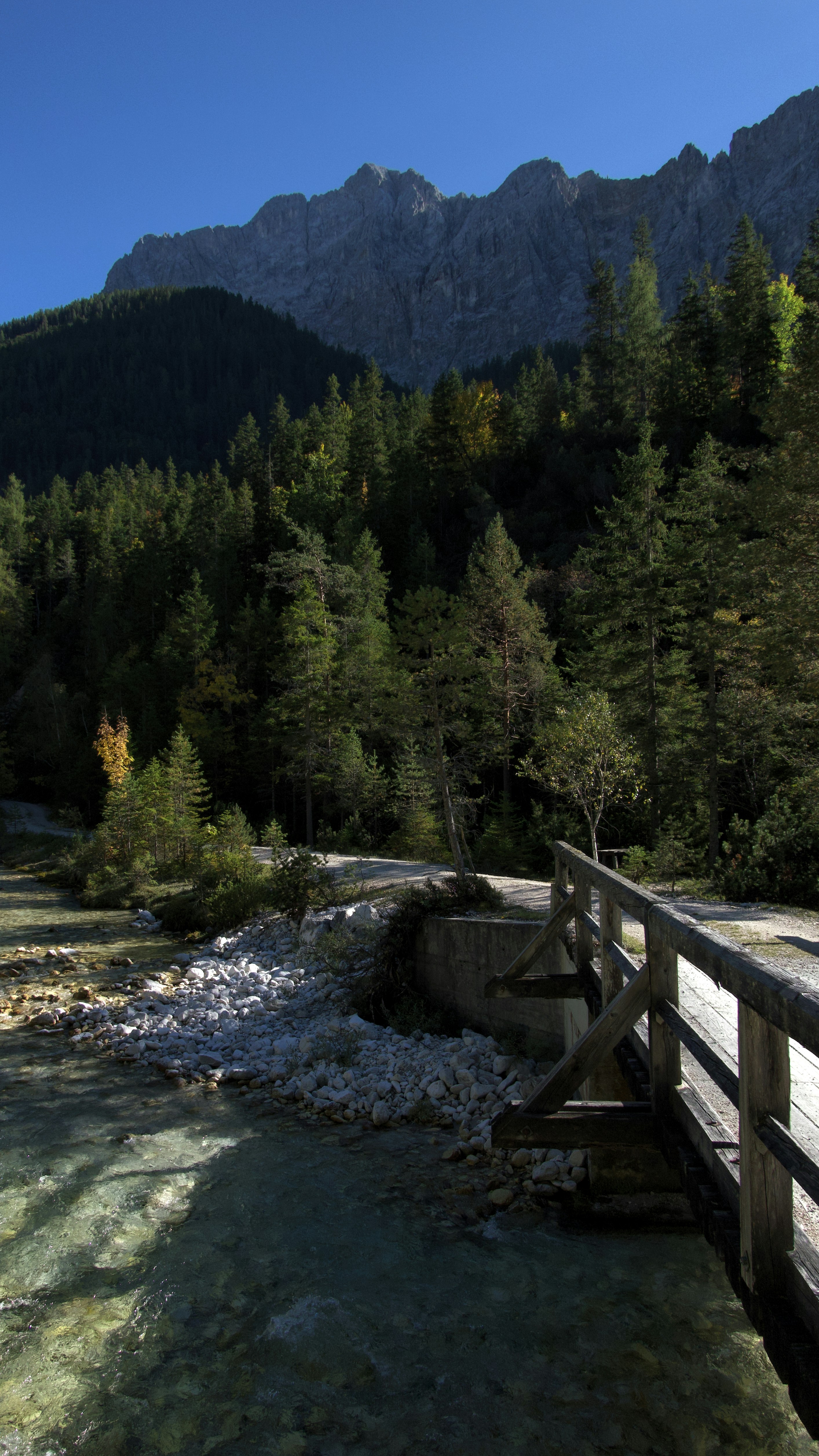 Wooden bridge crossing a clear stream, surrounded by lush trees and majestic mountains under a bright blue sky.