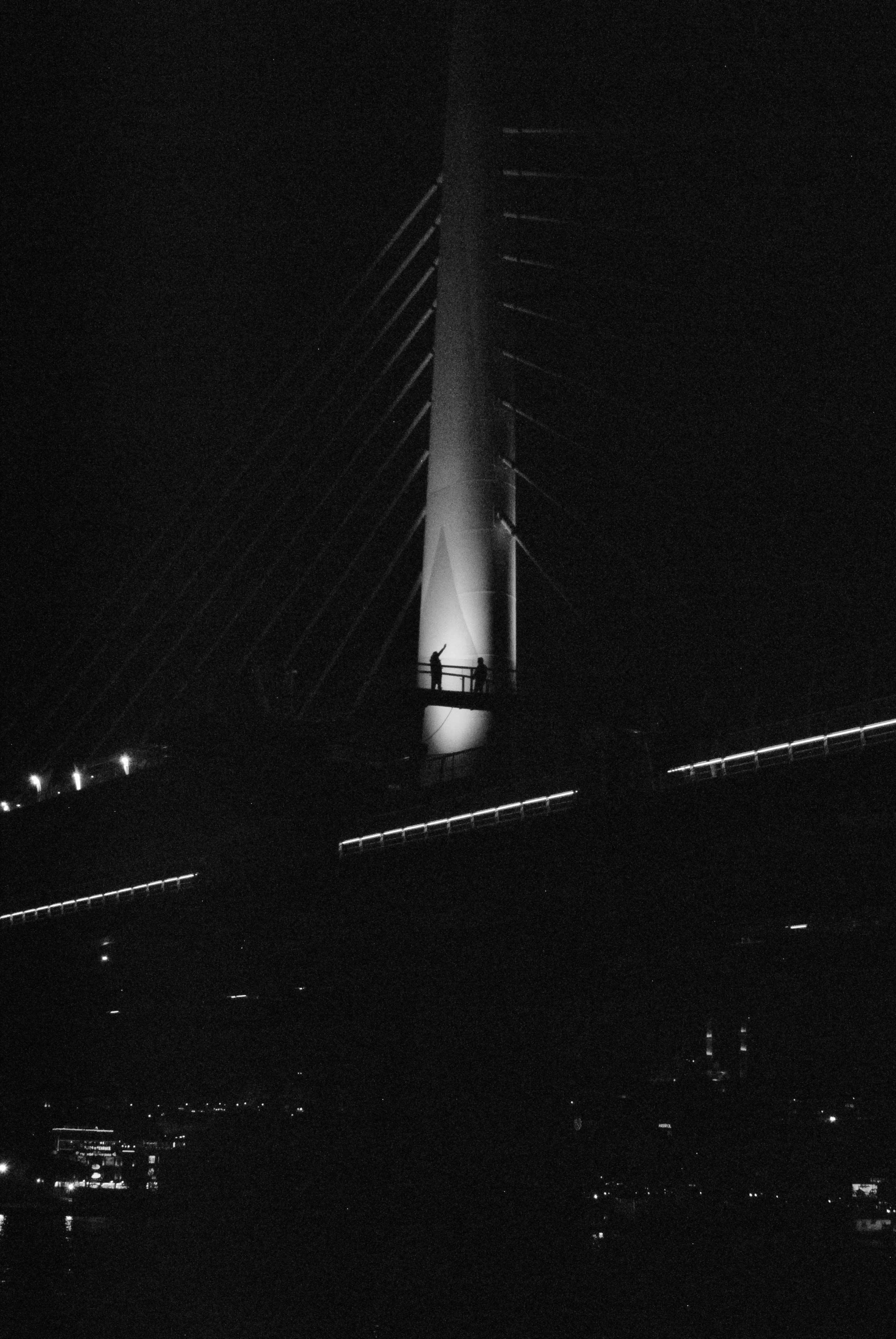 Night view of a modern cable-stayed bridge illuminated with lights, reflecting over the water. Minimal urban nightscape. | Illuminated bridge tower at night with city lights.