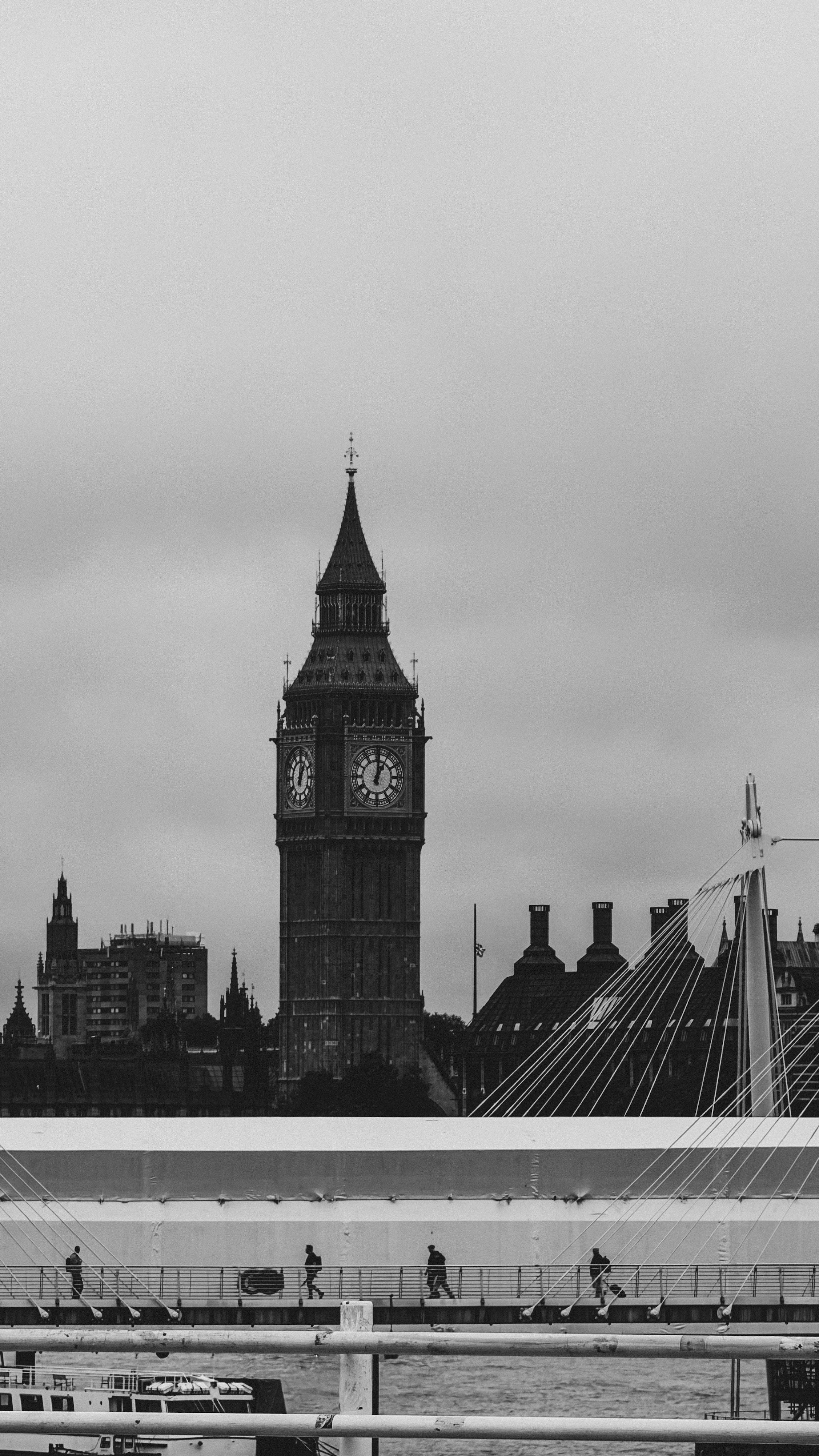 Elizabeth Tower stands against a grey sky, framed by Westminster rooftops and the Golden Jubilee Bridge. | Big ben and bridge on a cloudy day