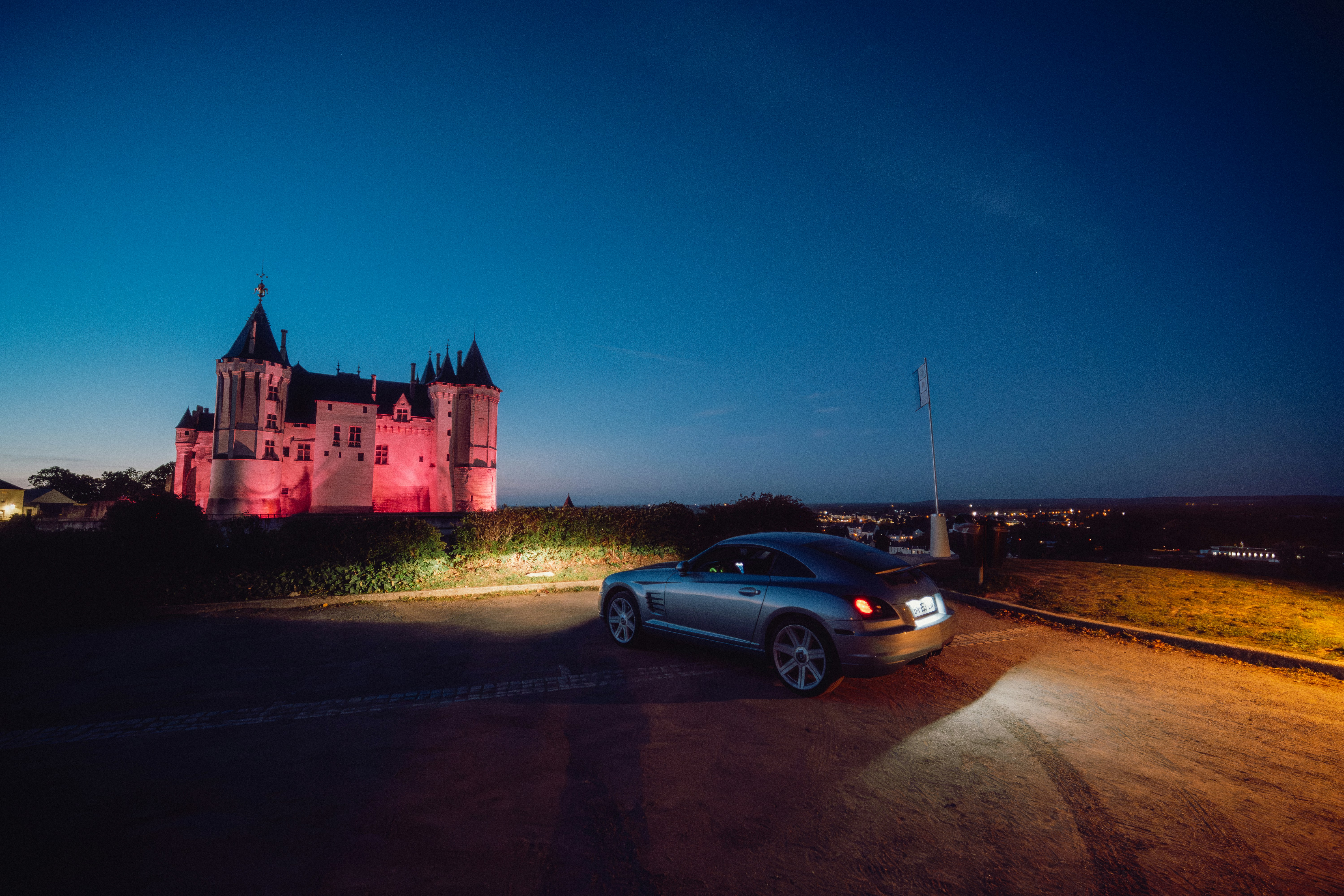 A sleek silver car parked near a historic castle illuminated in pink against a twilight sky.