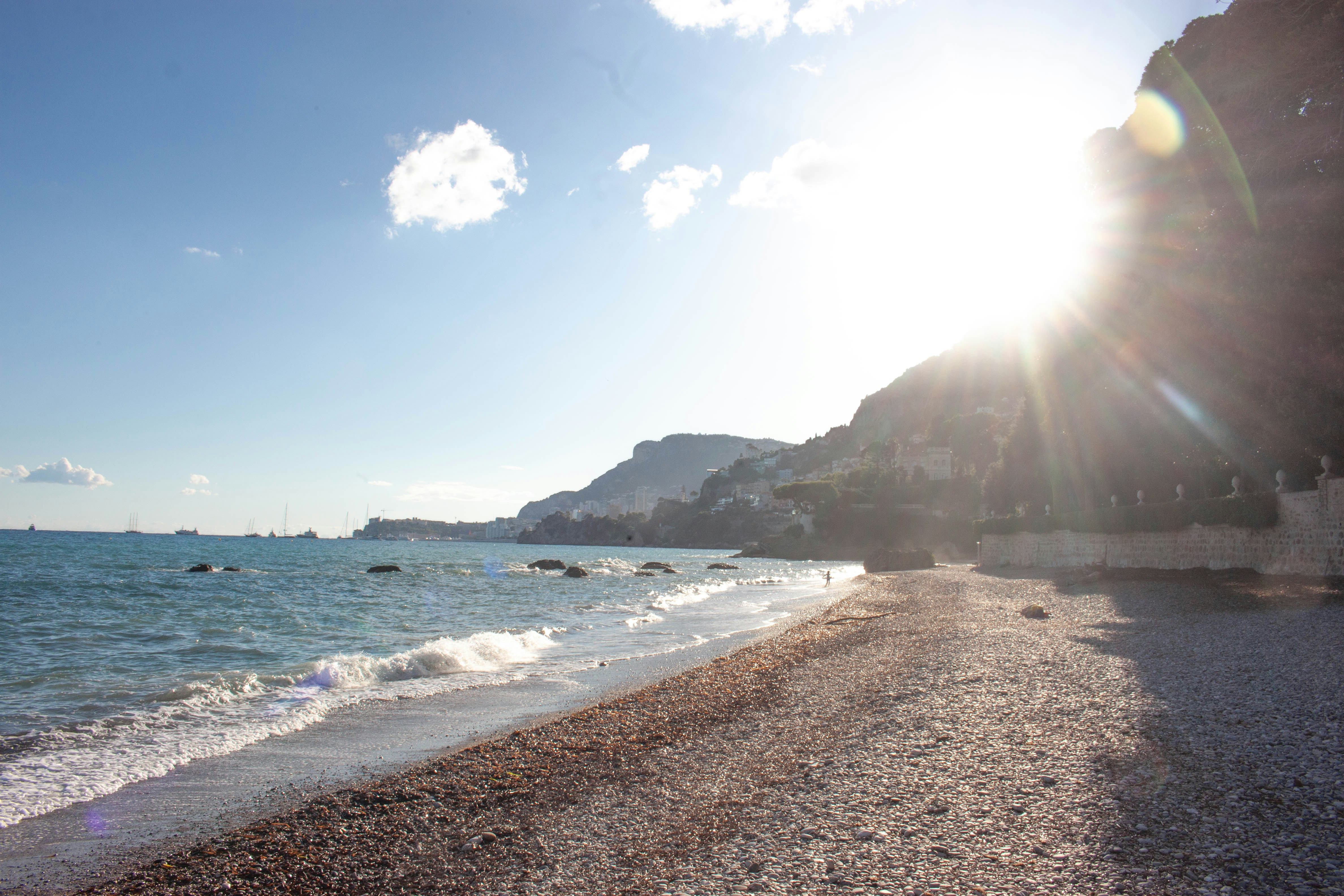 Golden sunlight glimmers over a tranquil beach, with pebbles lining the shore and distant mountains framing the scene.