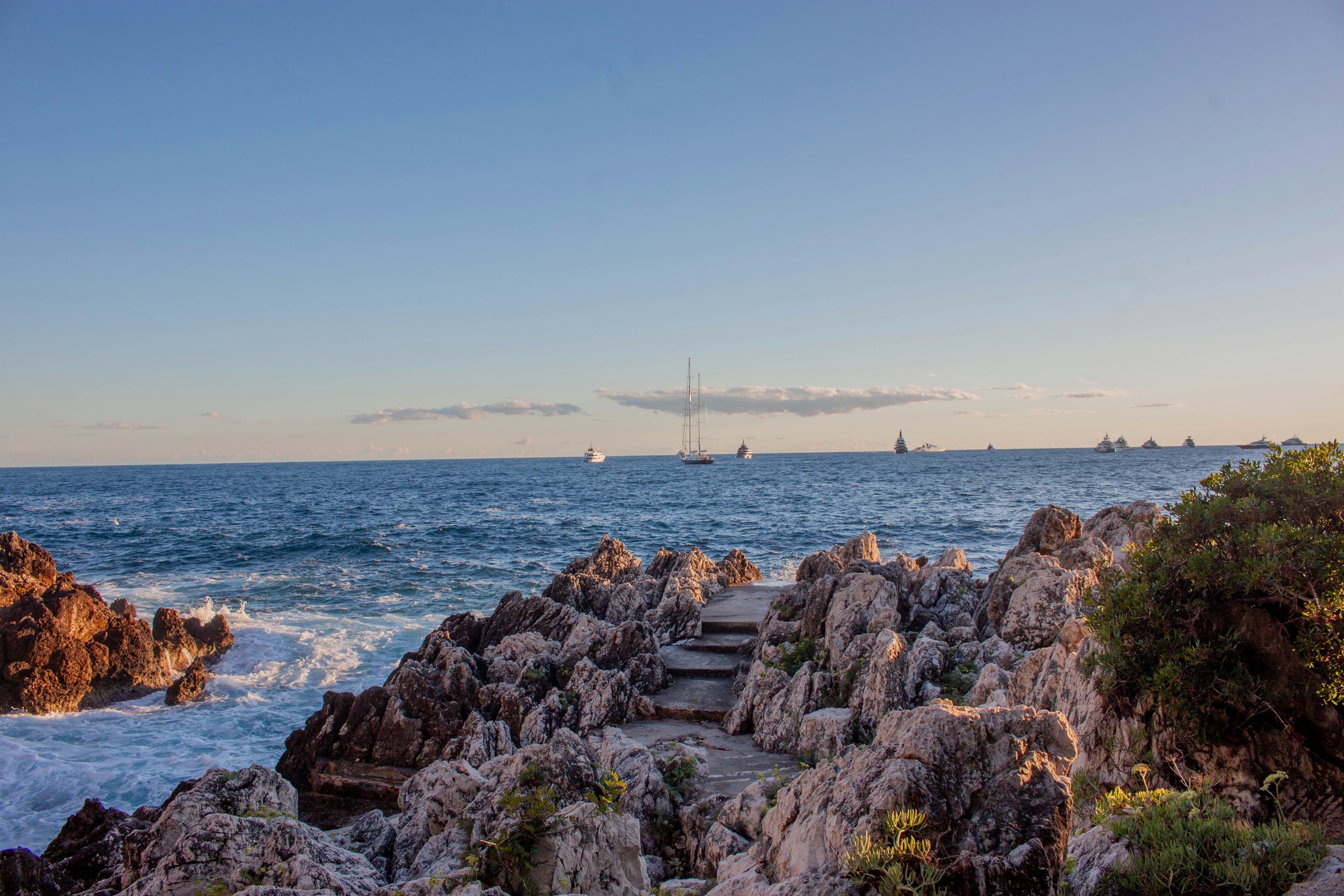 Rocky coastline leading to a serene ocean view with distant sailboats on the horizon.