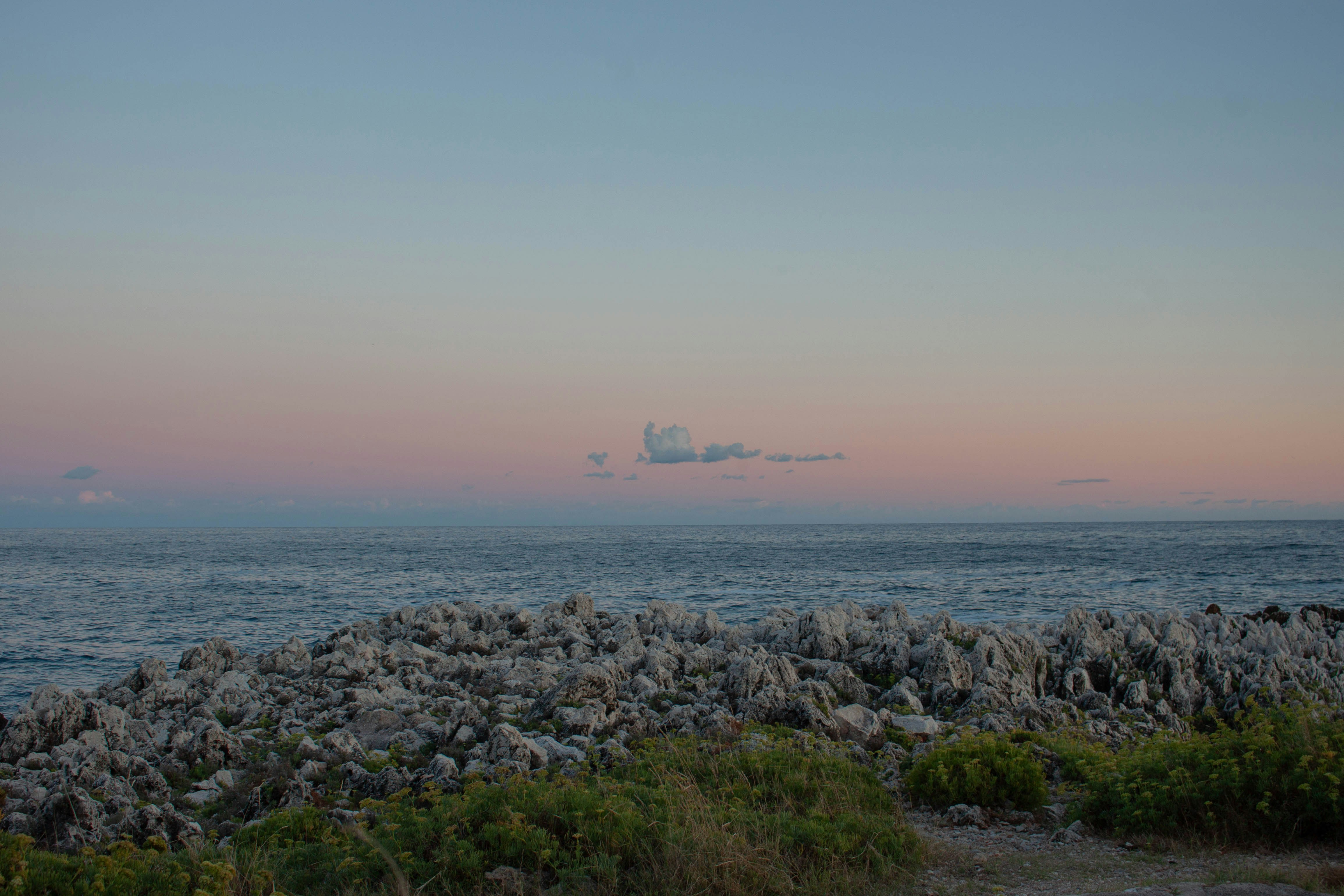 Rocky shoreline under a pastel sky at dusk, with gentle waves lapping at the stones. A solitary cloud drifts above the horizon.