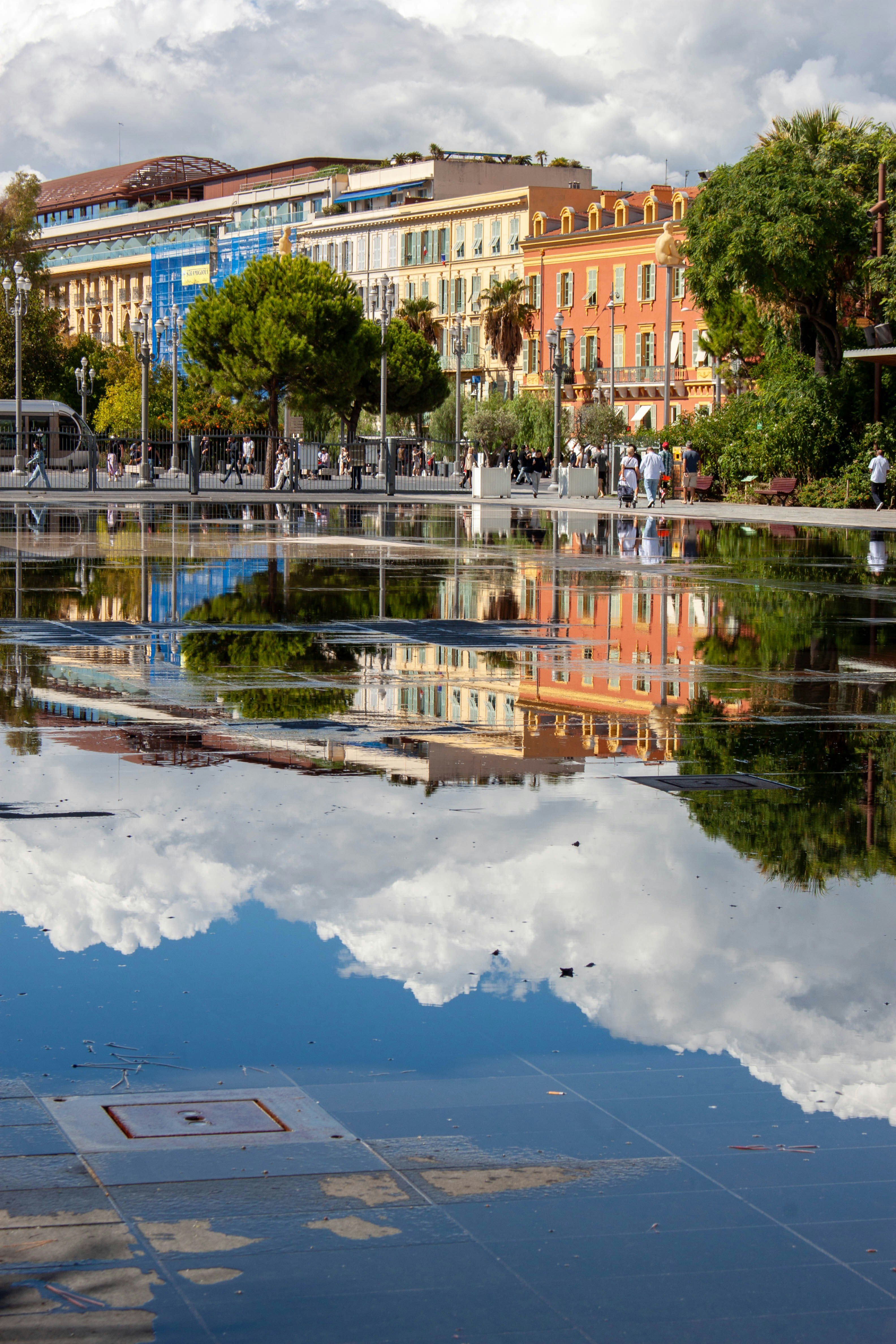 Buildings reflected in a wet plaza with clouds above.