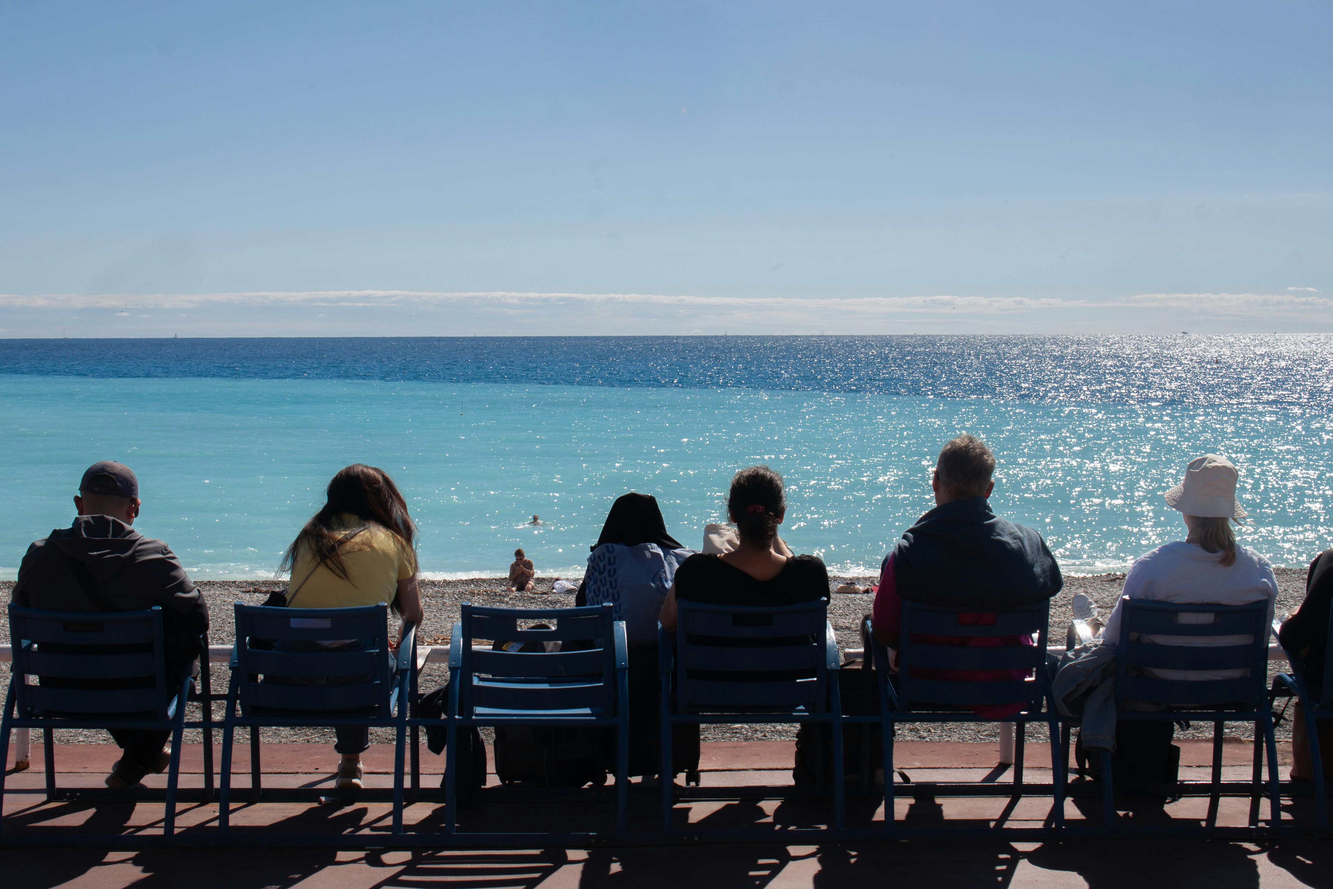 Group of individuals seated on blue chairs, gazing at a tranquil turquoise sea on a sunny day. The scene captures a peaceful coastal atmosphere.