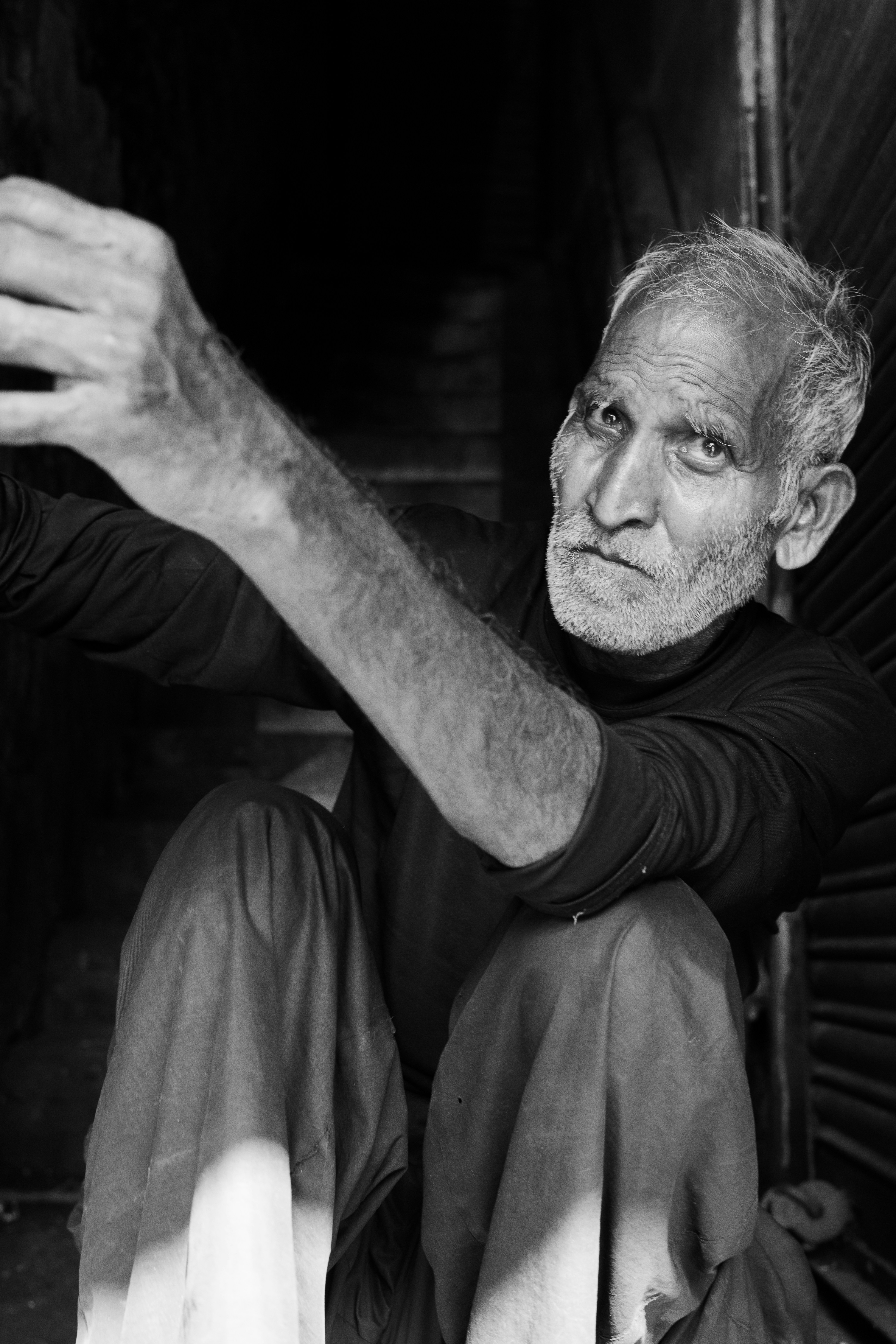 Elderly man seated in a dimly lit stairwell, extending his hand forward while showcasing a thoughtful expression in black and white.