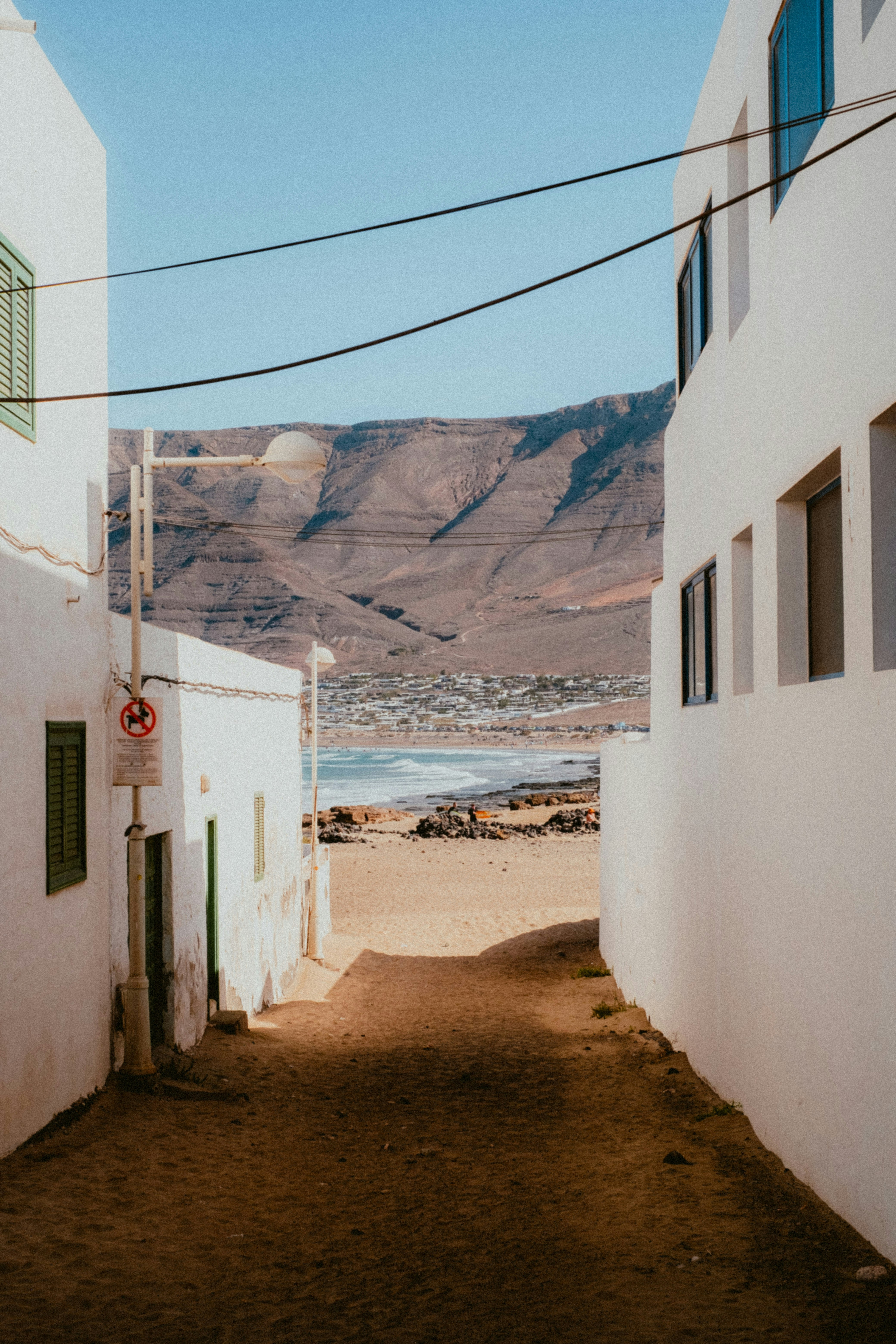Narrow sandy pathway between whitewashed buildings leading to a scenic coastal view with mountains in the background.