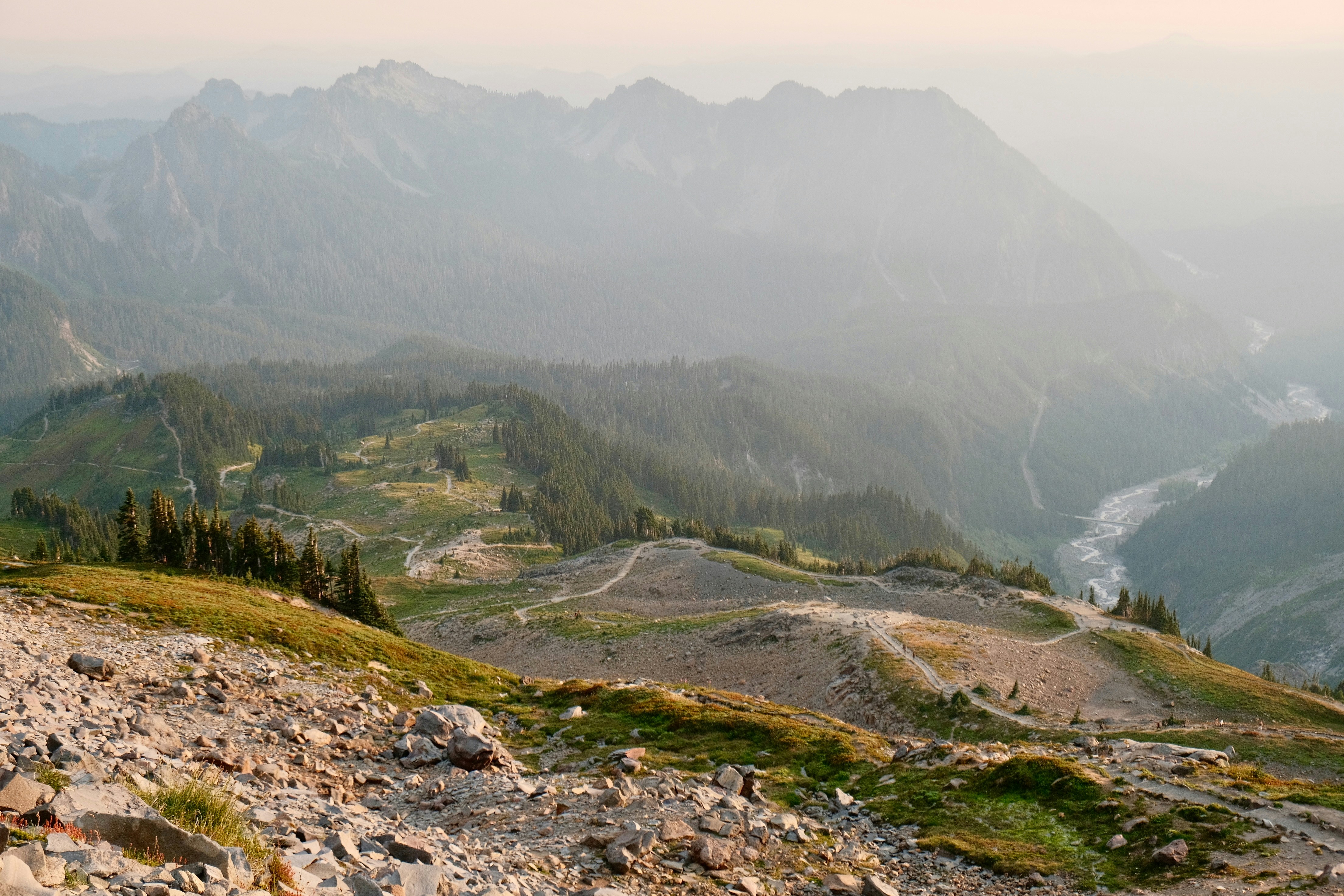 Expansive mountain vista showcasing rolling hills and a winding river, enveloped in a soft haze. The rugged terrain contrasts with the gentle light of dusk.