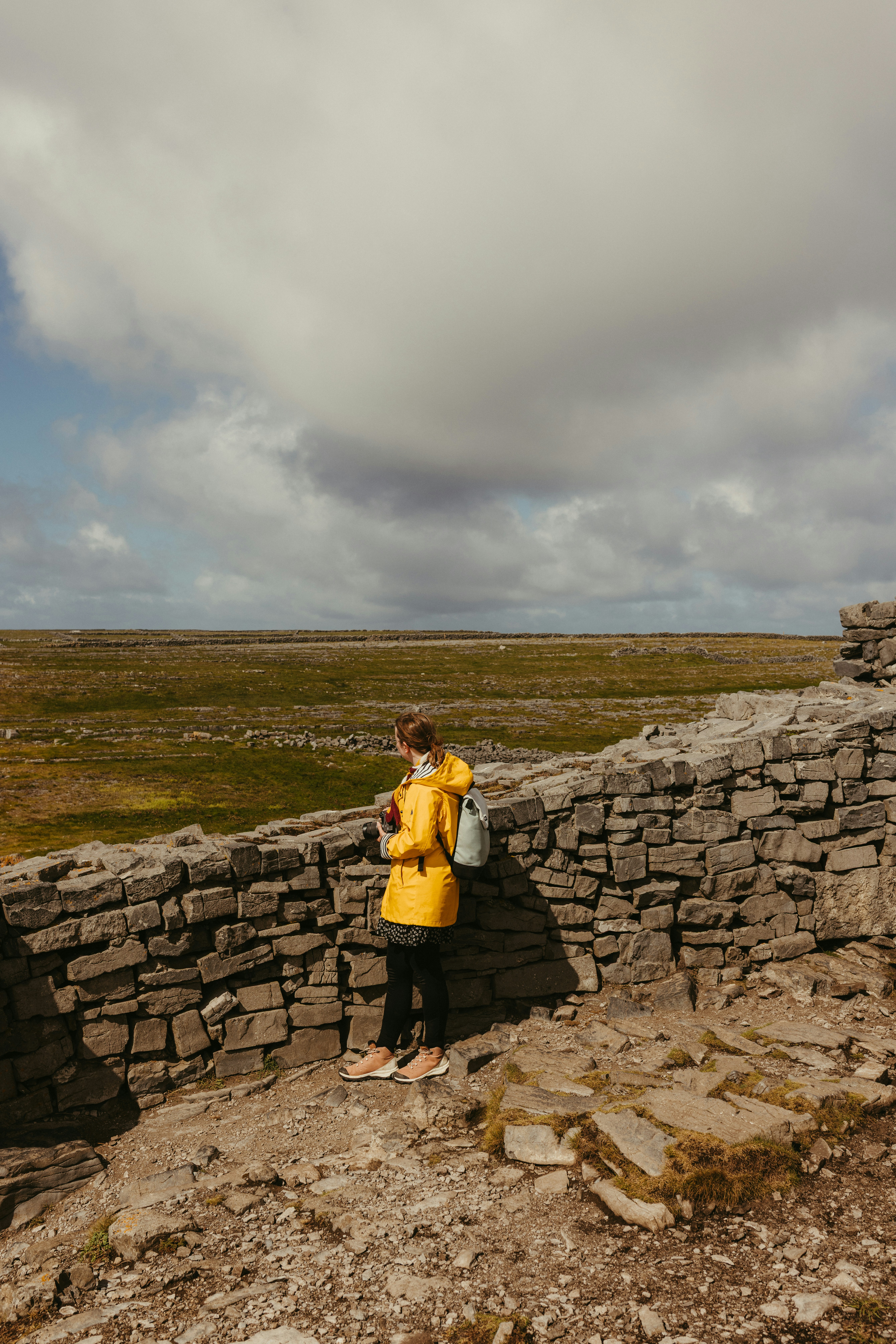 Person in a yellow raincoat stands beside a stone wall, gazing at the expansive landscape under a partly cloudy sky.