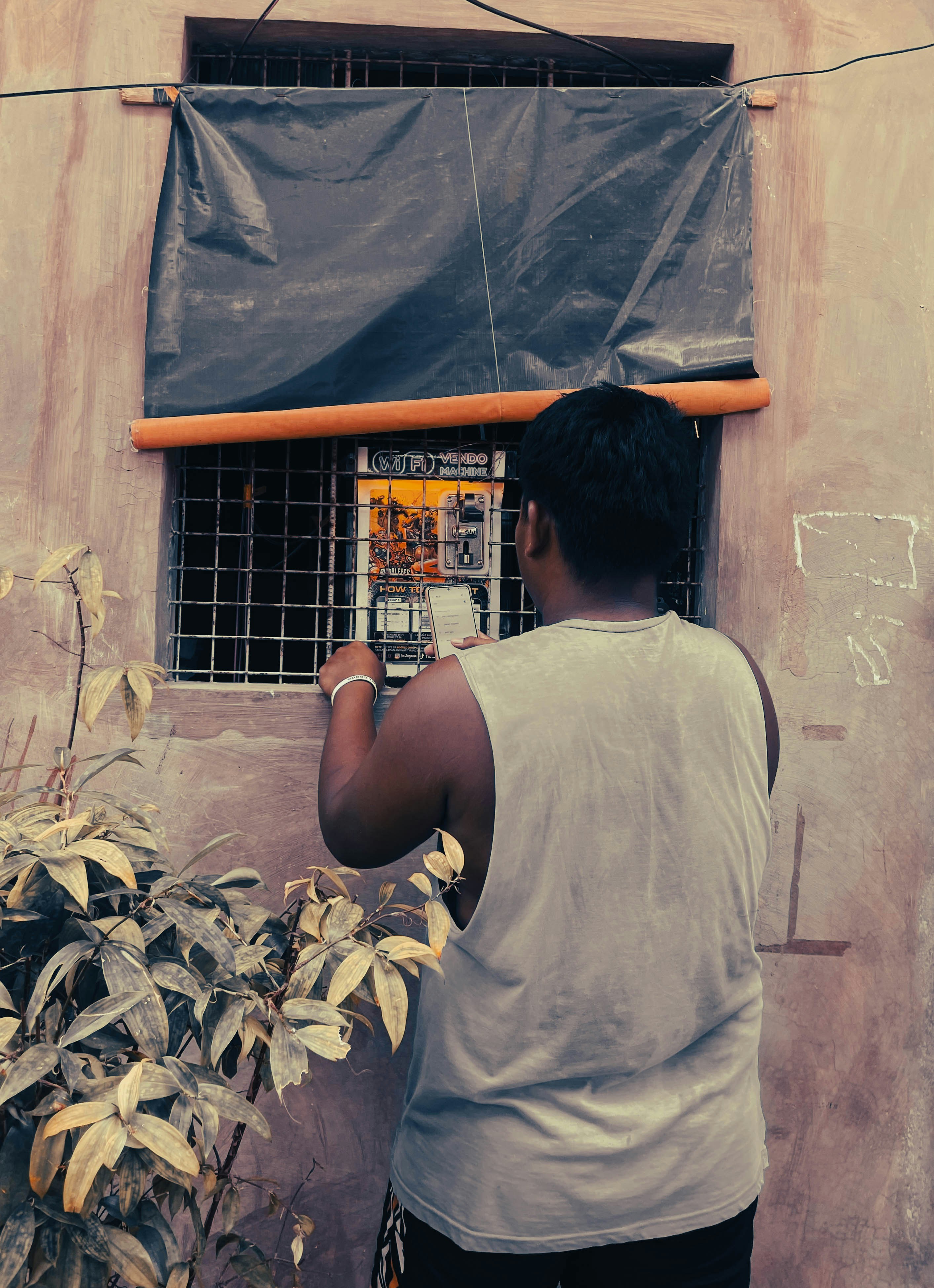 A person interacts with a vending machine through a barred window, surrounded by foliage. The scene captures a moment of daily life.