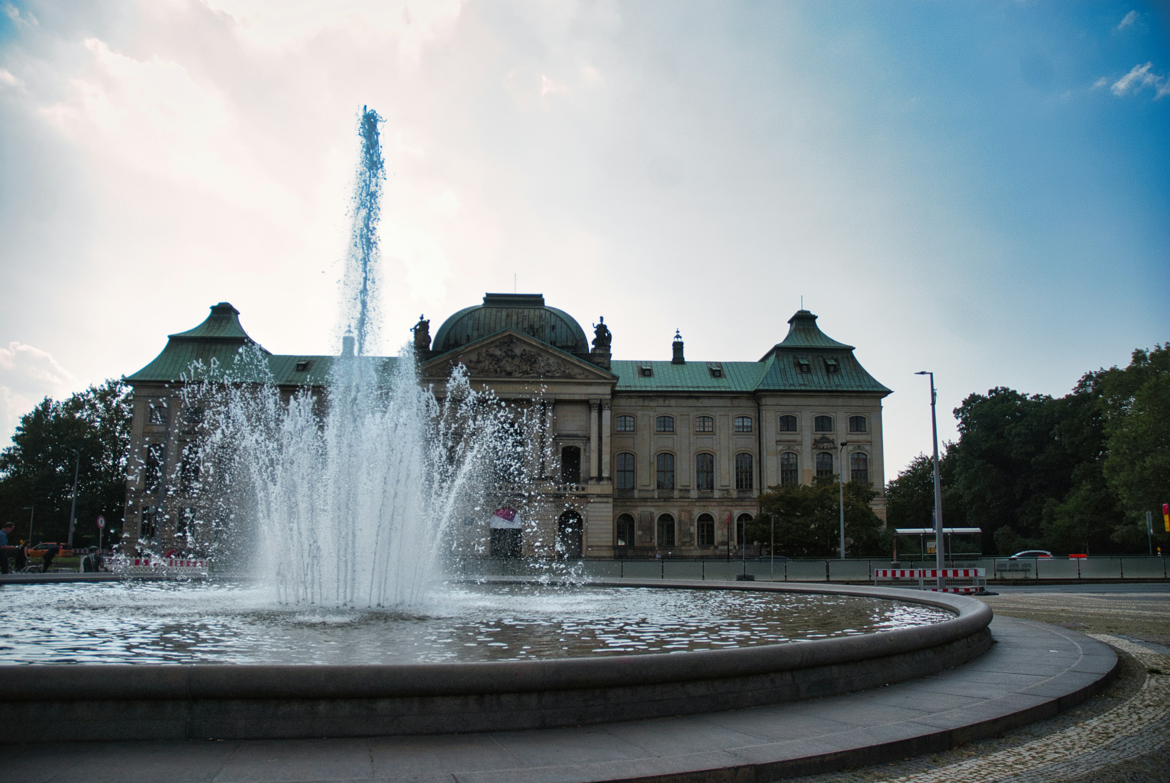 Majestic fountain spraying water in front of a historic building with green domed roofs and a cloudy sky.