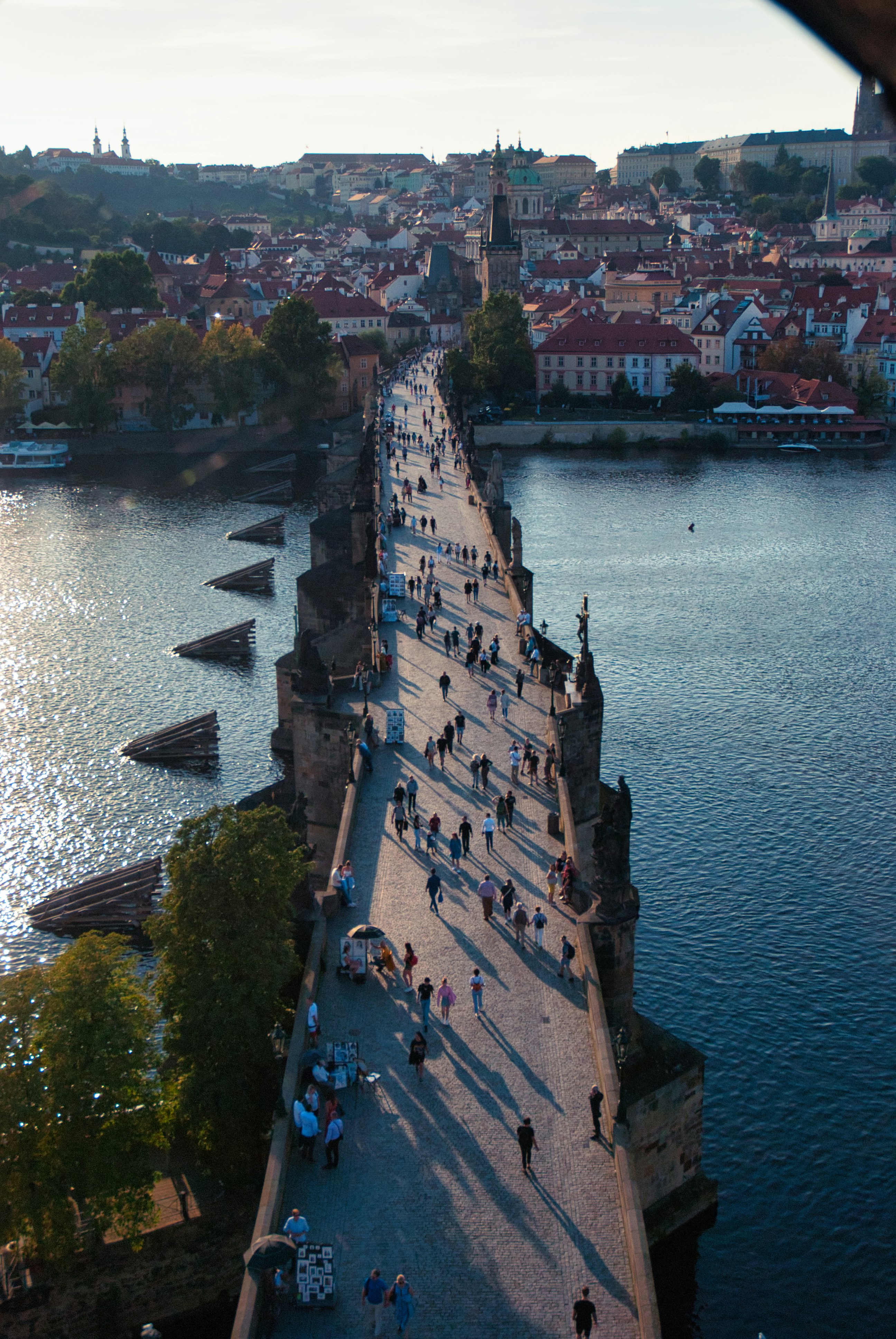 Pedestrians stroll along the historic Charles Bridge, framed by the scenic backdrop of Prague's architecture and river. Shadows stretch across the stone path.