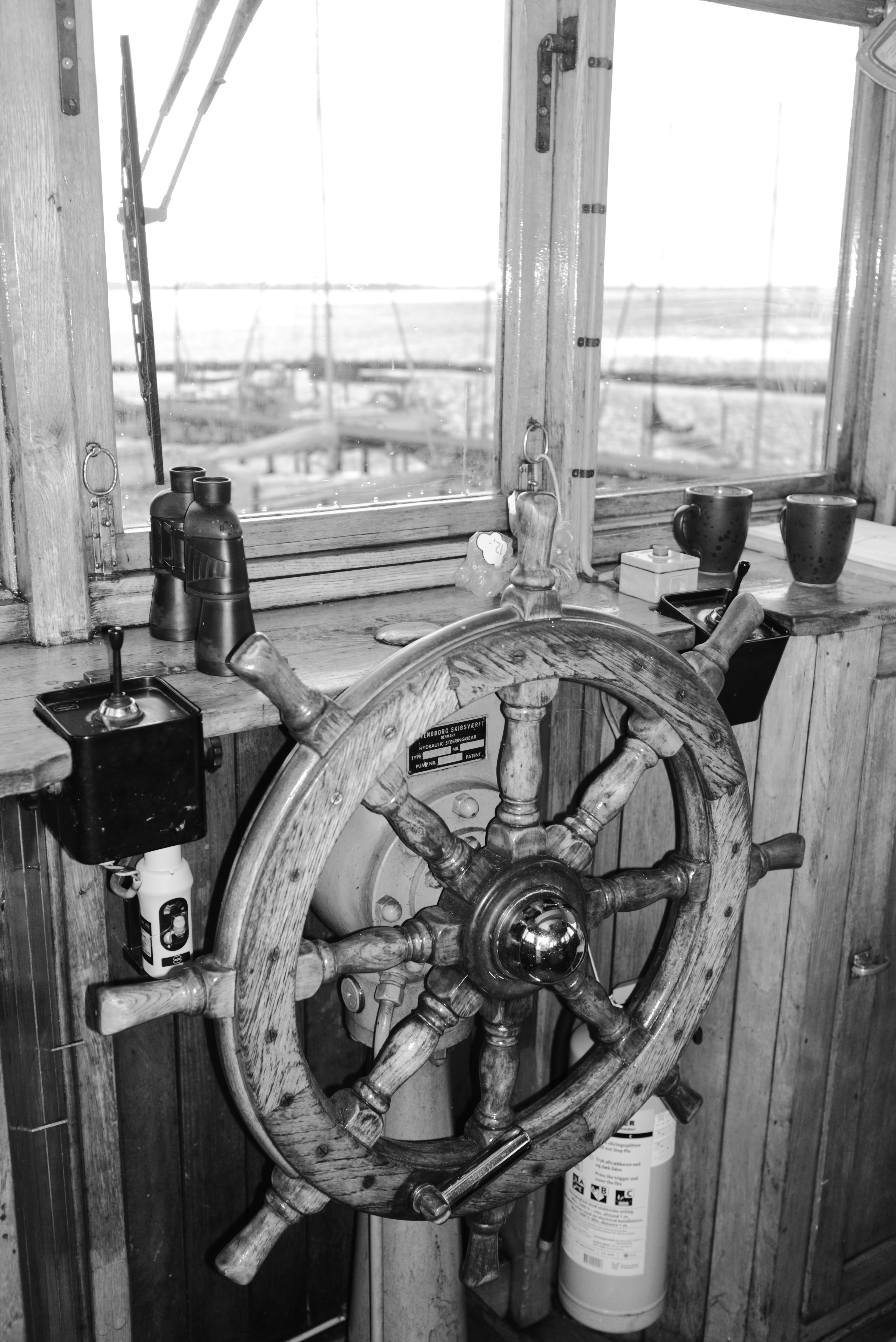 A vintage ship's wheel set against a backdrop of nautical equipment and a distant view of the water. The scene evokes a sense of maritime history.
