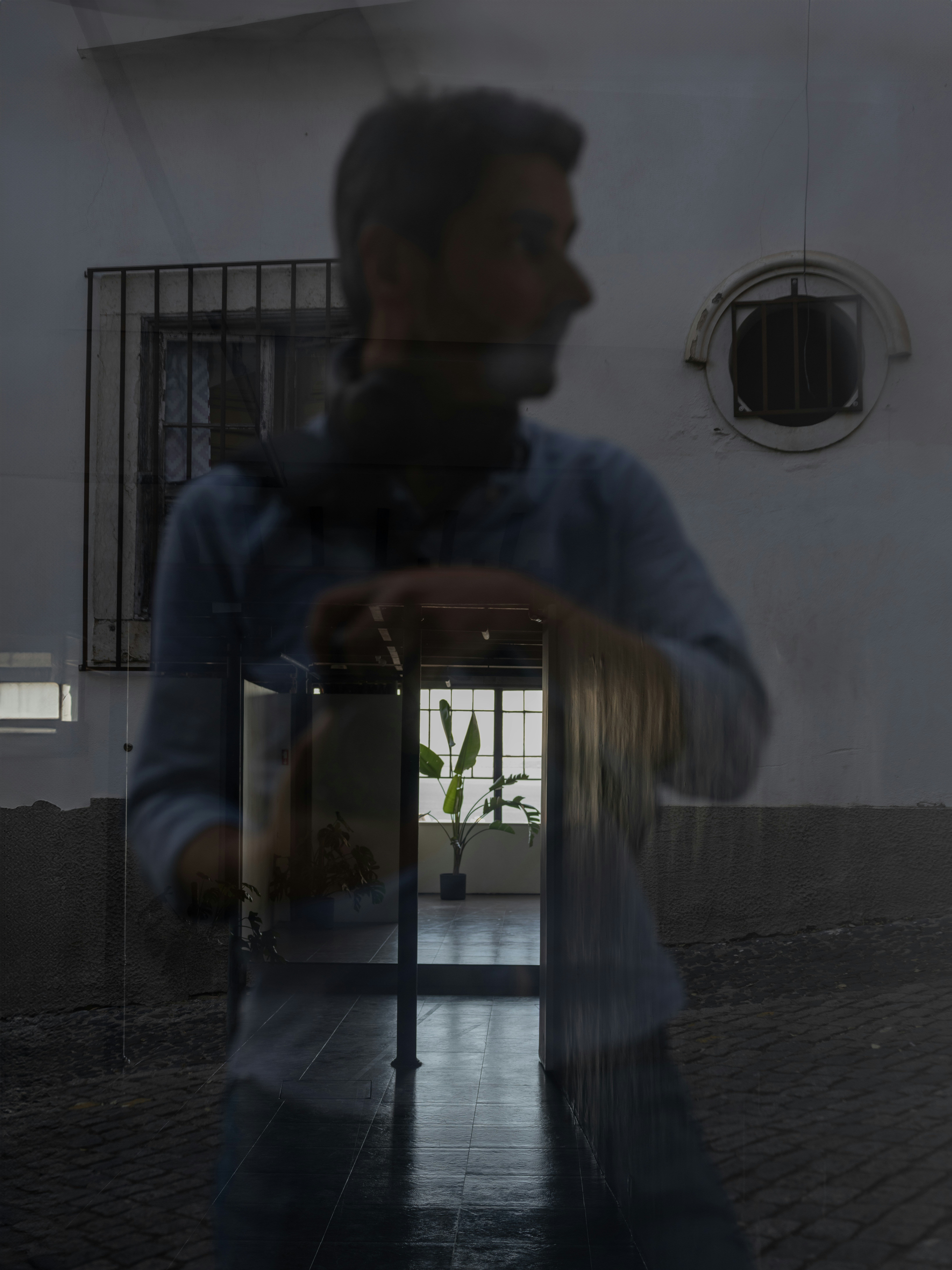 When I was born, there were already names waiting for me, and a few that had been forgotten. The stories of who I was moved softly through the rooms, carried in recipes and gestures. «You look just like him,» they’d say, or «you have that same stubborn way of standing.» Years passed, and some versions of me became solid, while others turned to dust. I learned to wear faces the way people wear seasons: one for belonging, one for being. A few never fit quite right. | Man reflected in window with plant in room