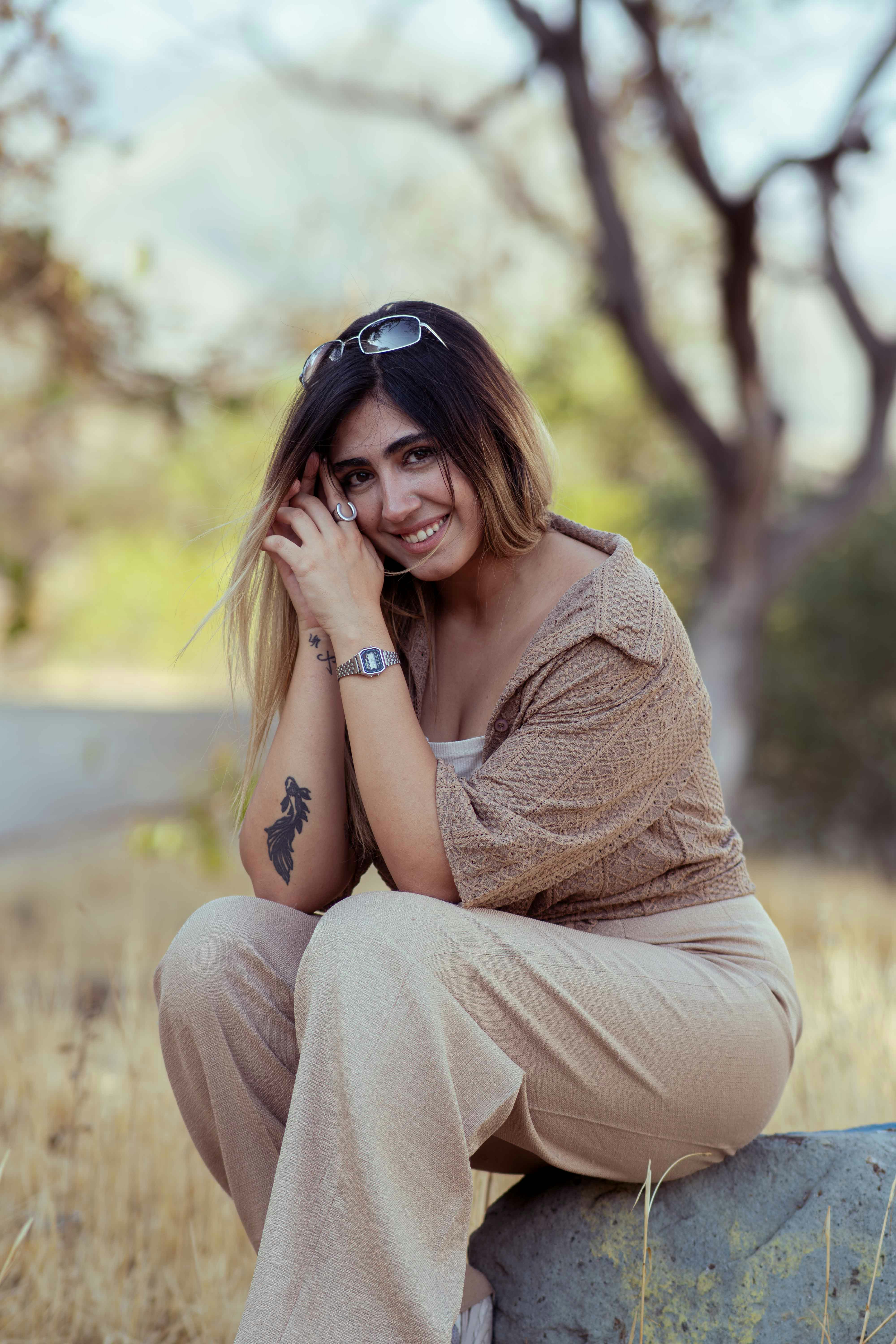 Woman sitting on a rock in a sunlit outdoor setting, exuding a relaxed vibe with a gentle smile and casual attire.