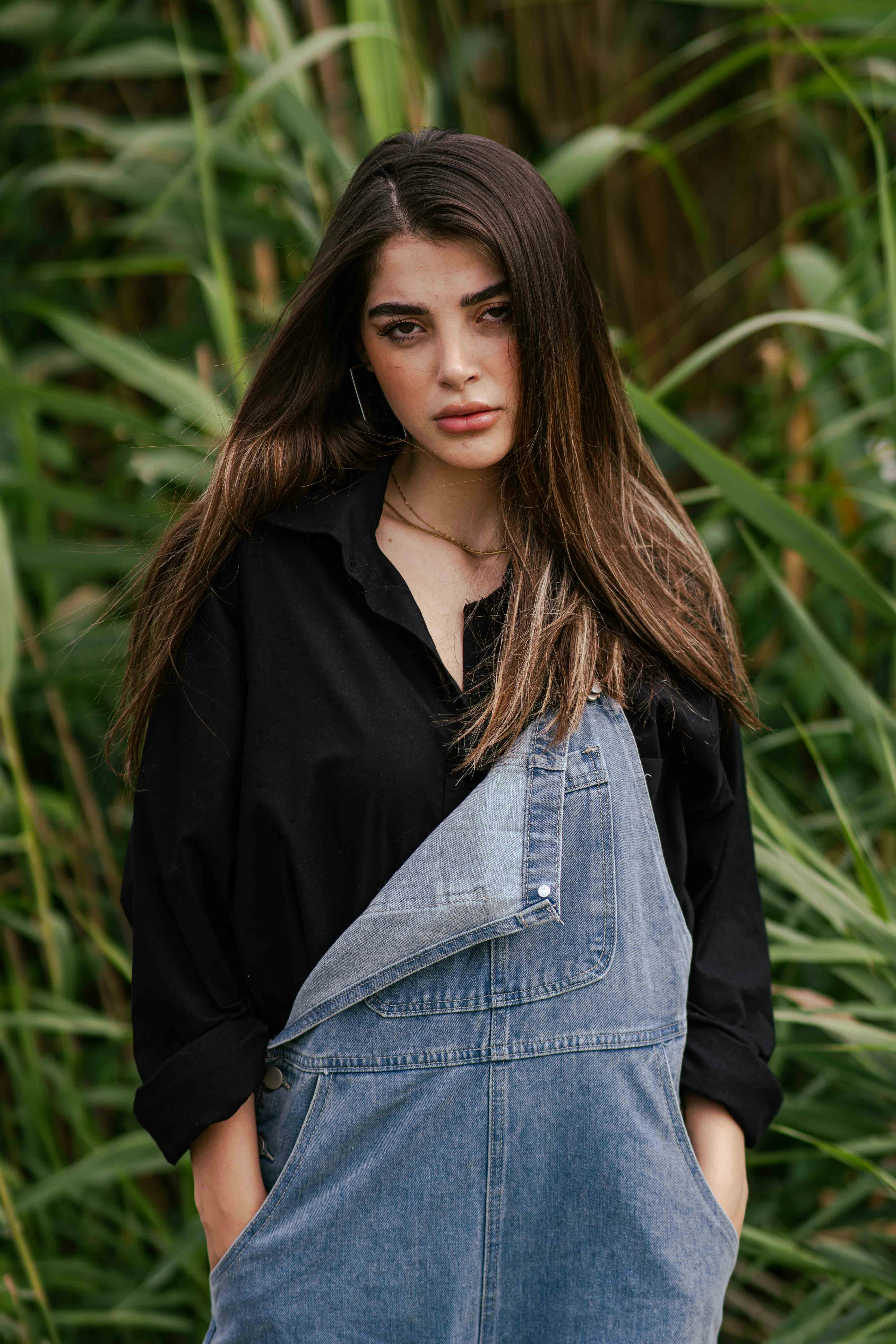 A young woman with long hair poses confidently in a black shirt and denim overalls, surrounded by lush green foliage.