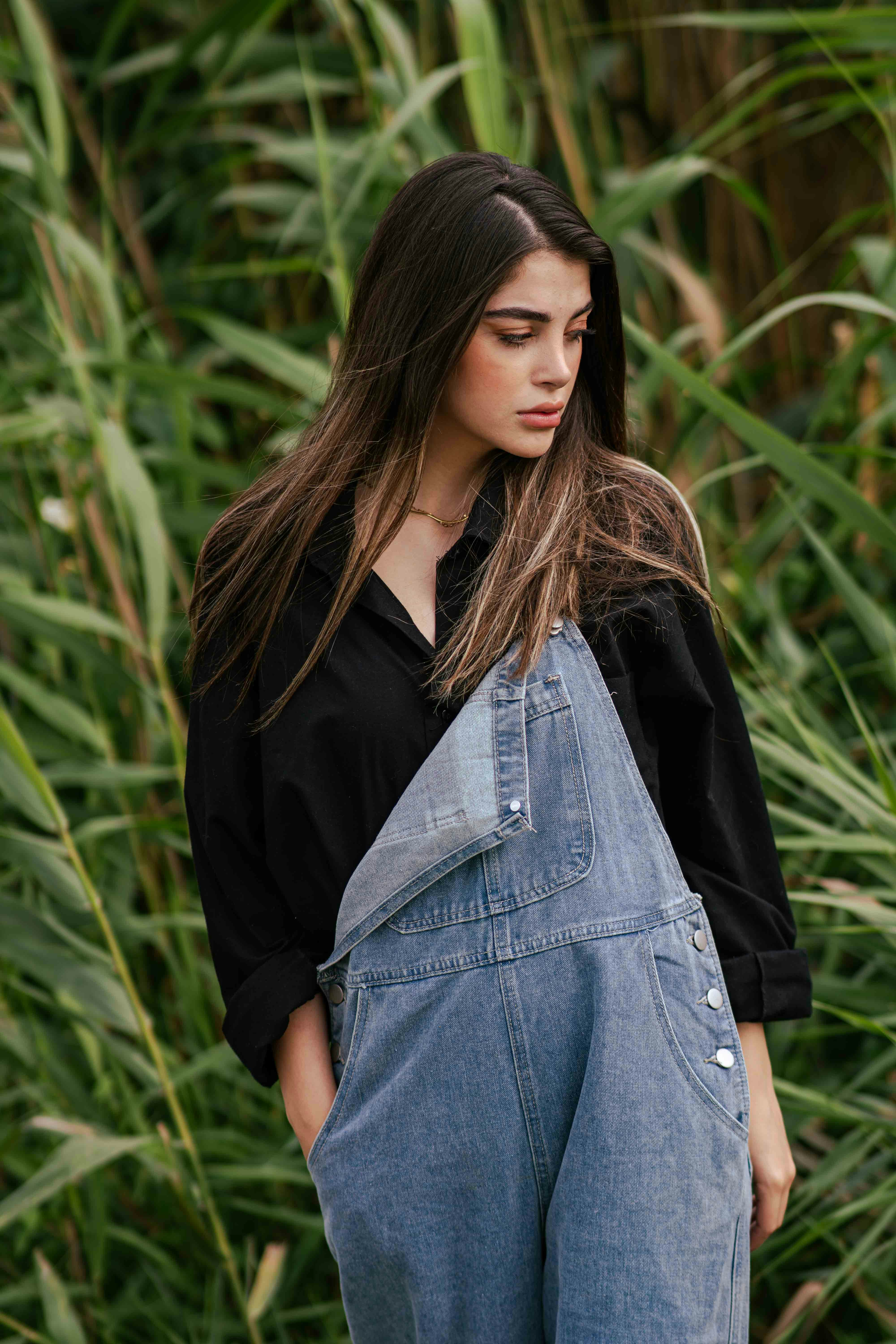 Young woman in a black shirt and denim overalls stands amidst tall green foliage, exuding a calm and introspective vibe.