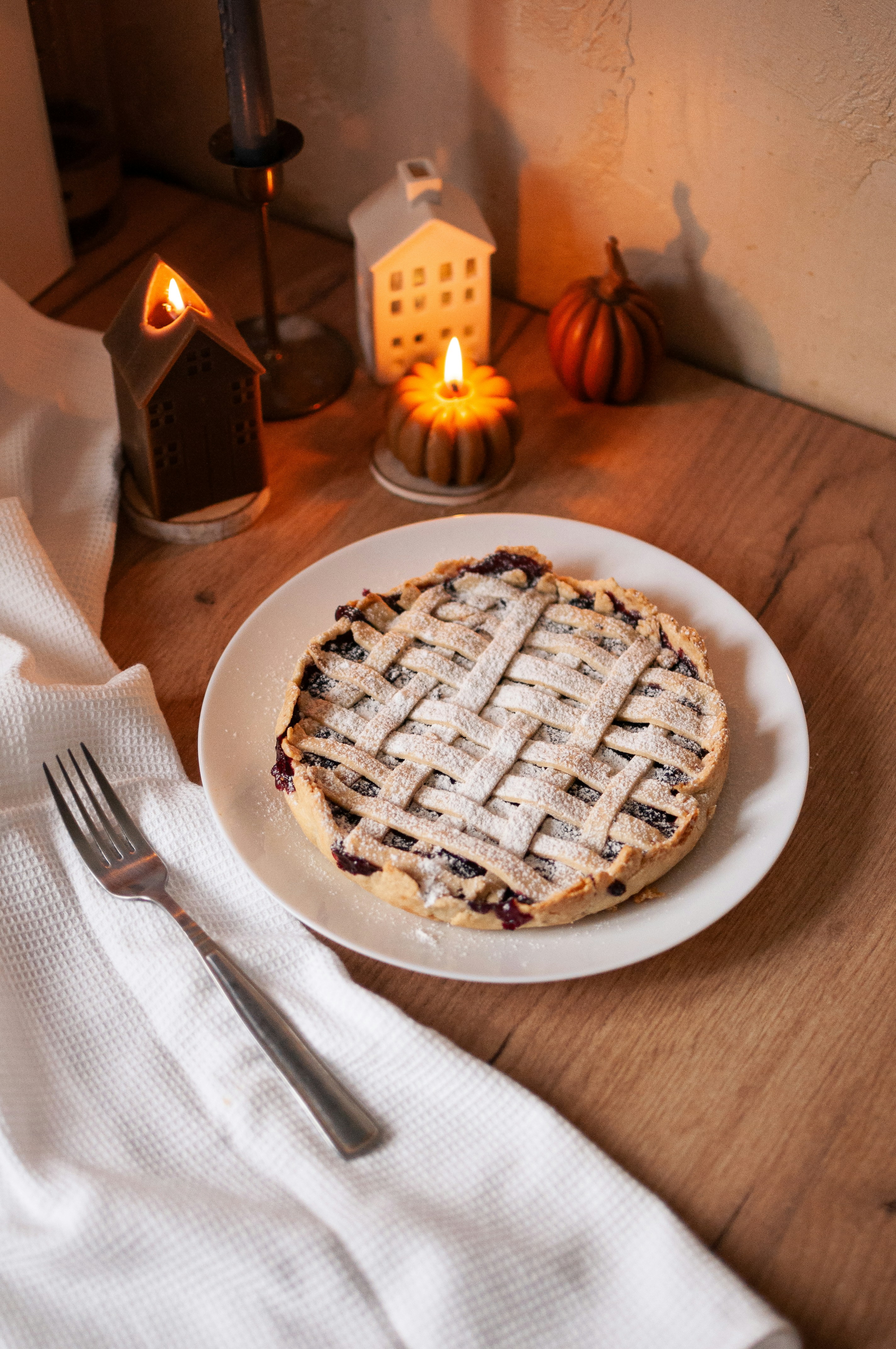 A freshly baked pie dusted with powdered sugar sits on a white plate, surrounded by decorative candles and small pumpkins, evoking a warm autumn atmosphere.