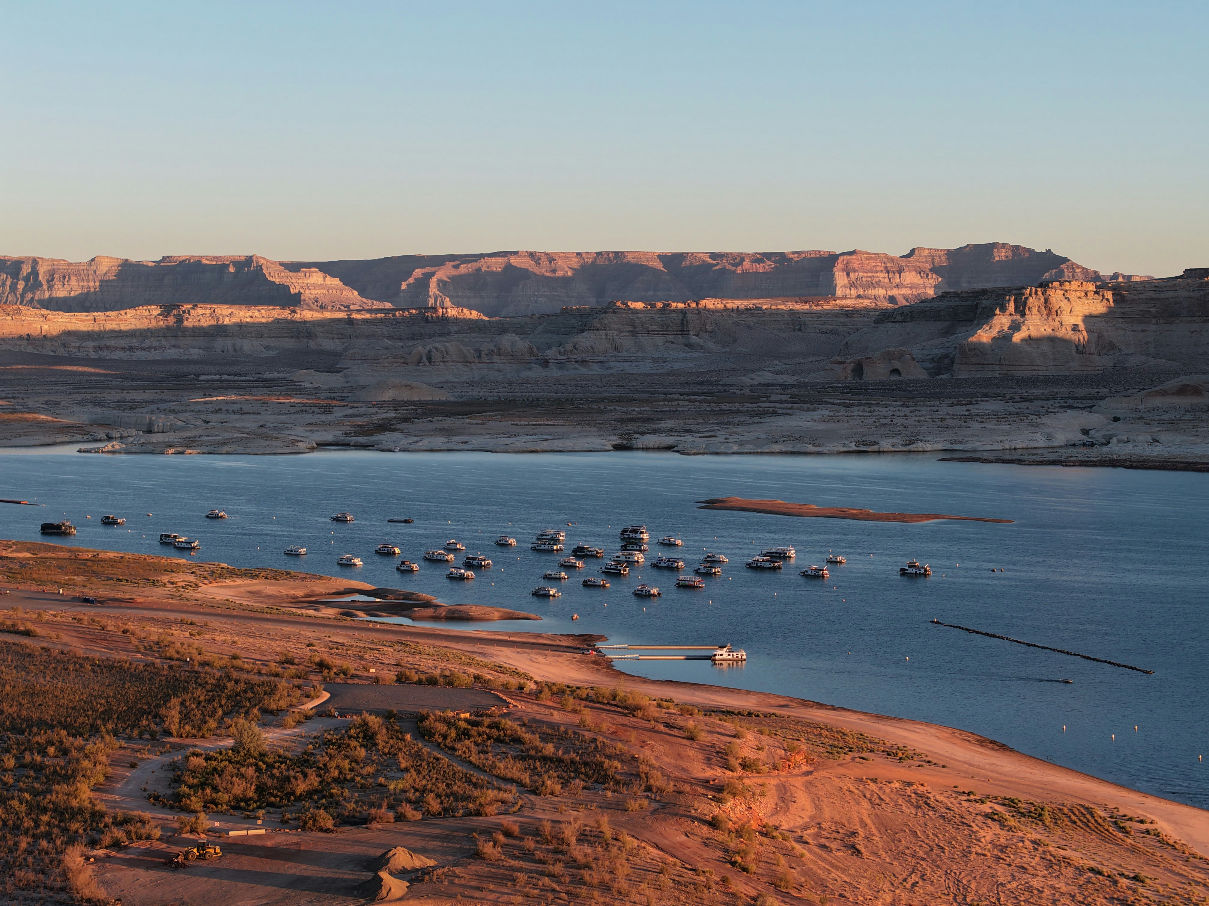 A tranquil scene featuring boats anchored in a calm lake, surrounded by rugged cliffs at sunset. The warm hues of the setting sun accentuate the landscape.