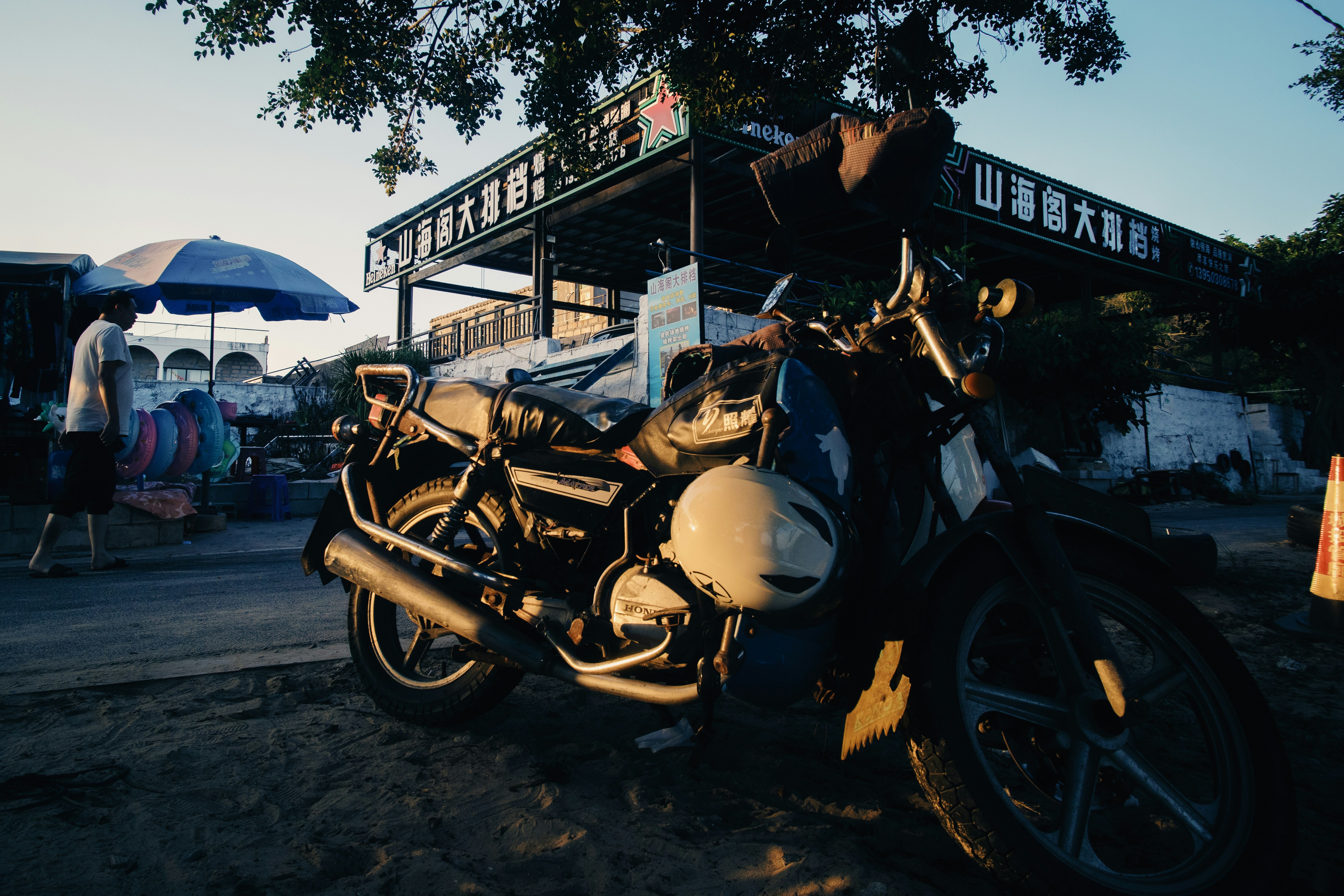 Vintage motorcycle parked under a tree near a bustling market, capturing the essence of local culture and adventure.