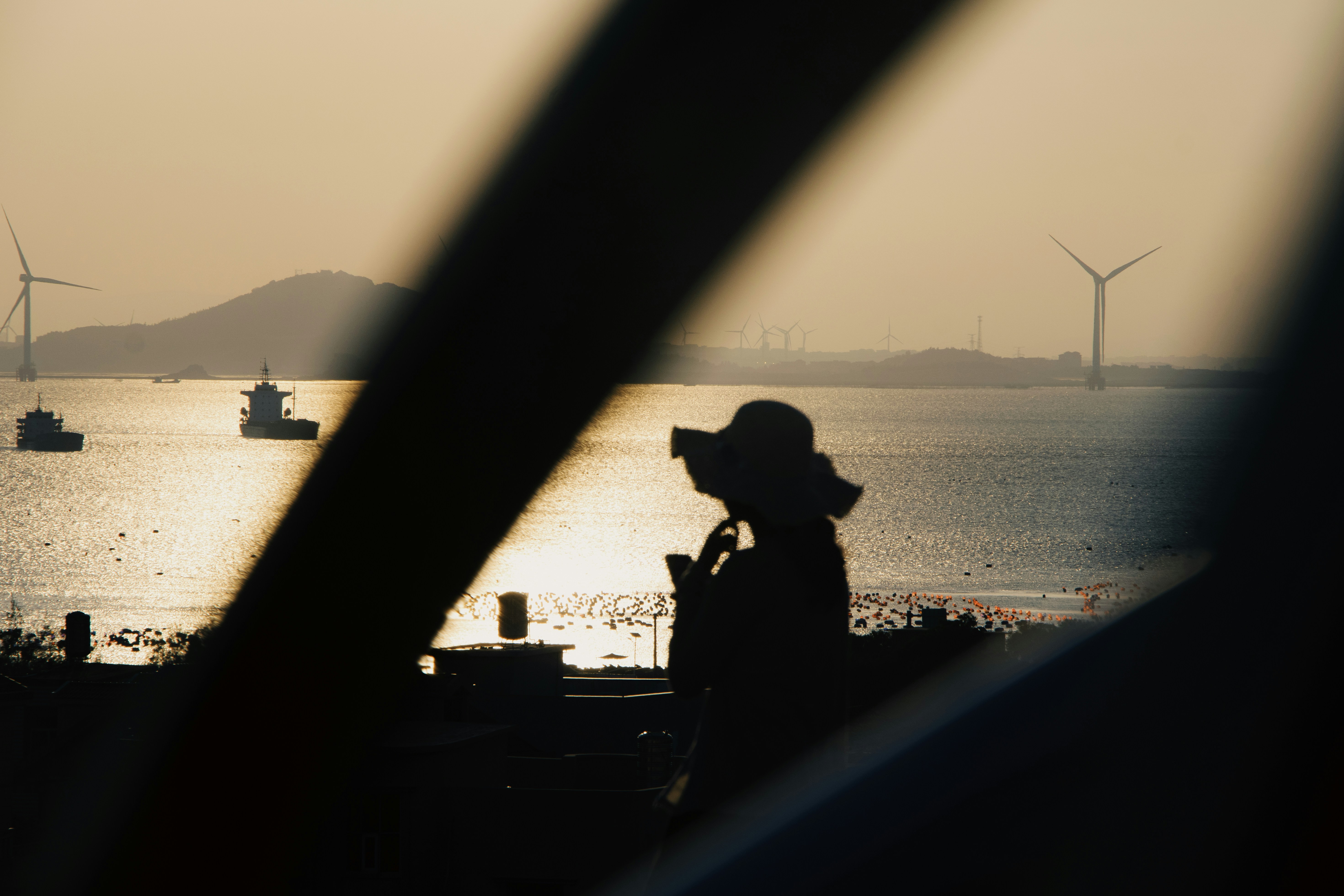 A silhouette of a woman with a wide-brimmed hat stands against a shimmering sea at sunset, framed by the outlines of nearby structures. Wind turbines dot the horizon.