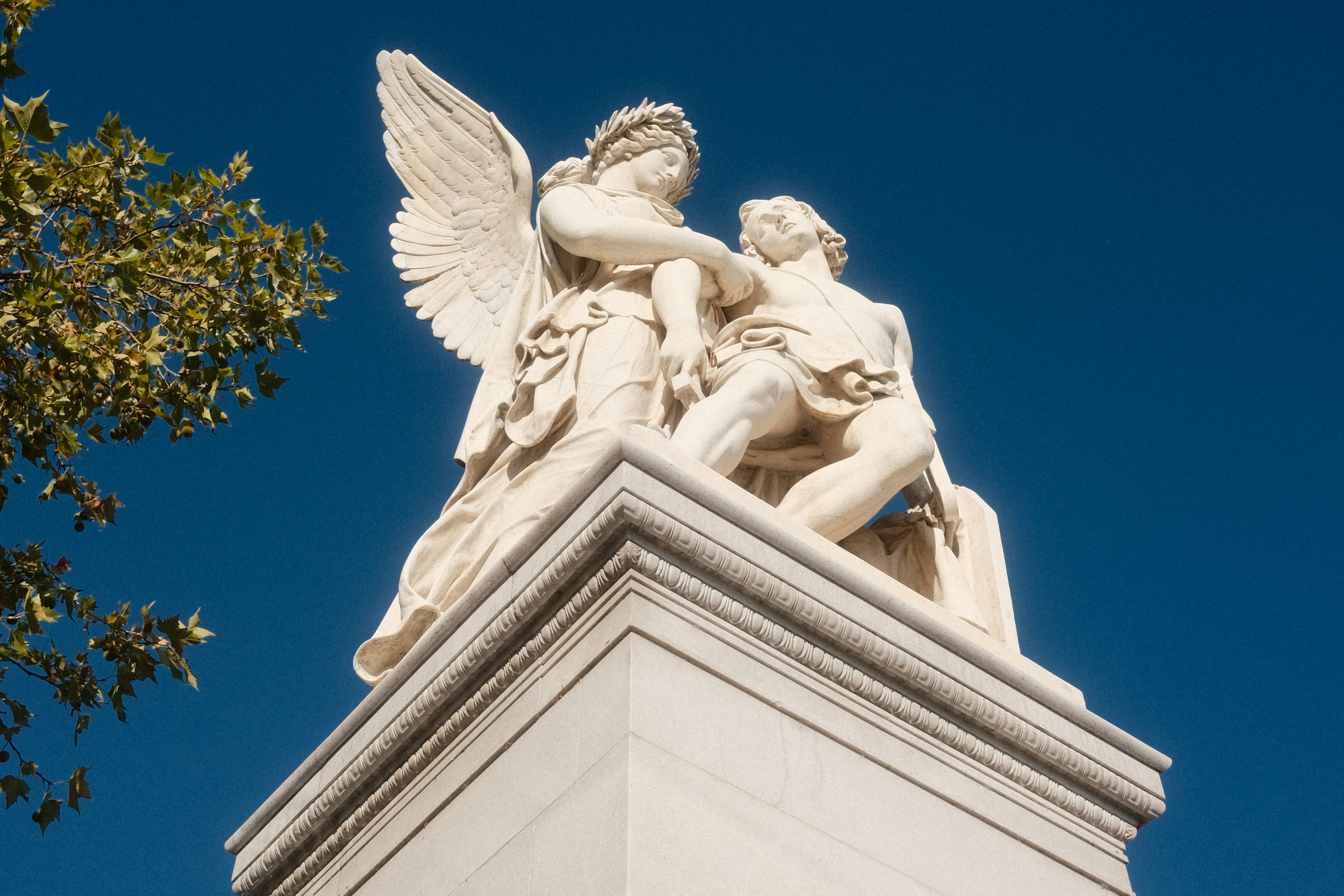 A marble statue depicting an angelic figure interacting with a young man, set against a clear blue sky. The intricate details of the sculpture are highlighted by the sunlight.
