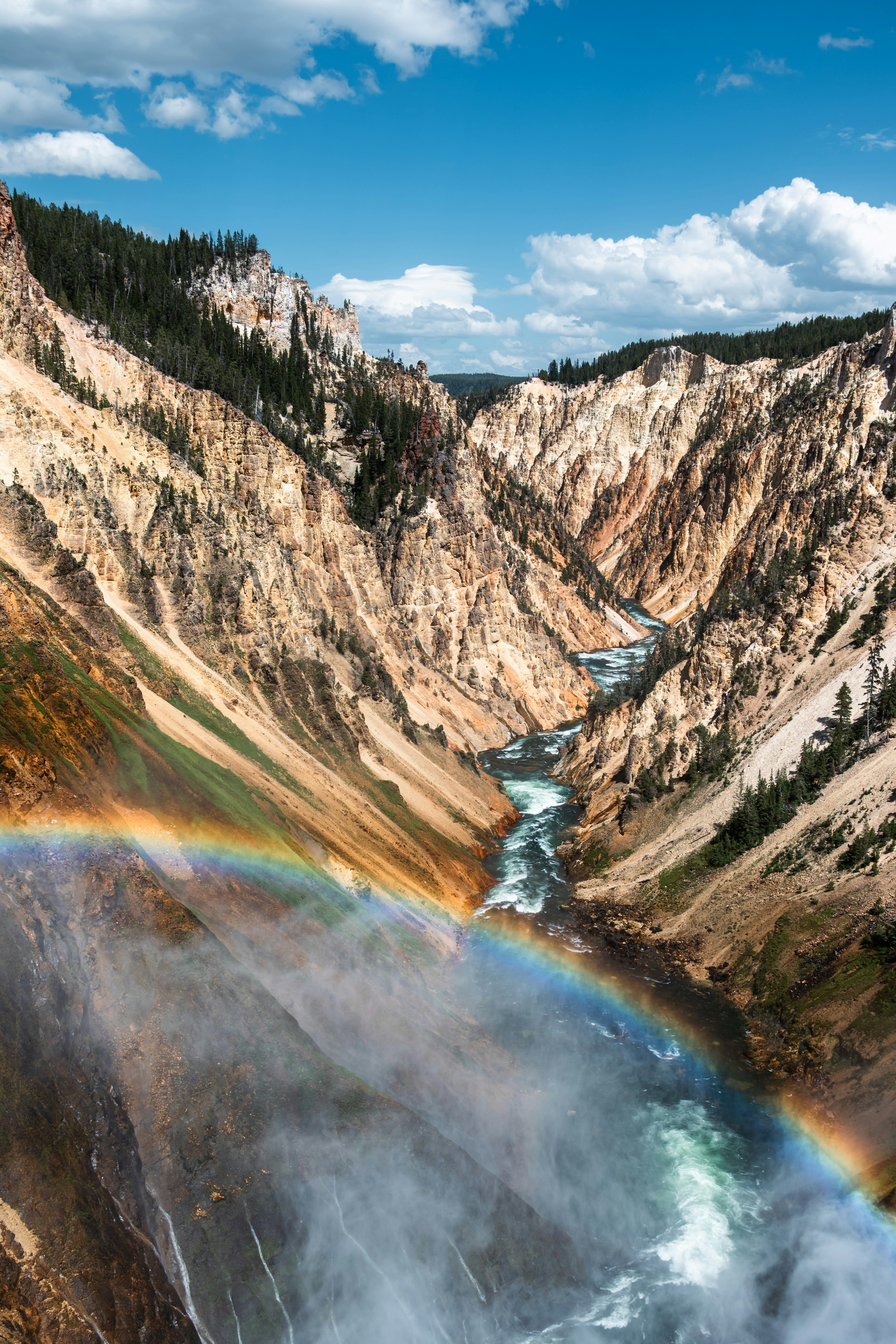 A vibrant rainbow arches over a cascading waterfall, with rugged cliffs framing a winding river below. The scene captures the dynamic interplay of water and light.