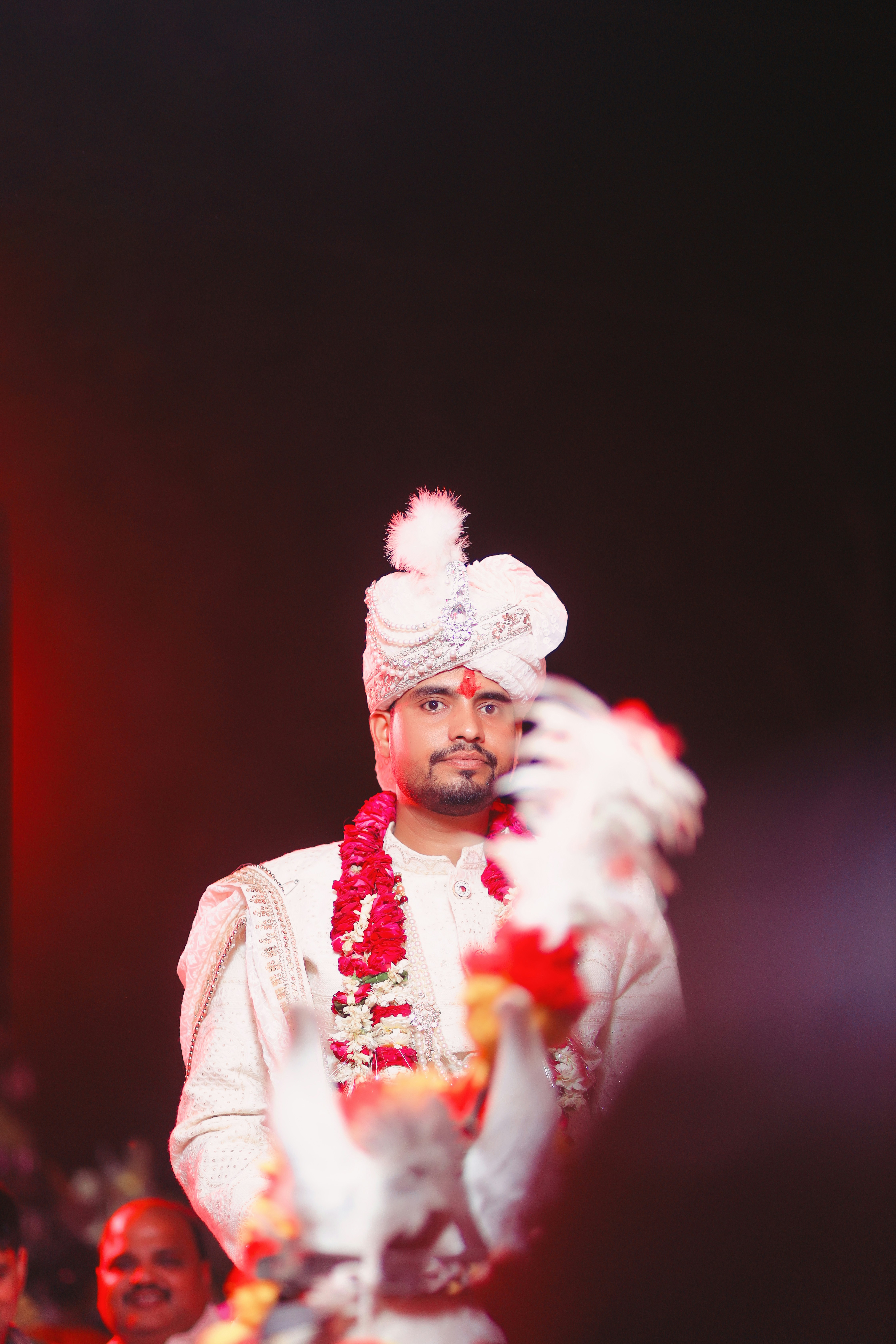 A groom in ornate traditional attire adorned with floral garlands, standing poised amidst a festive atmosphere.
