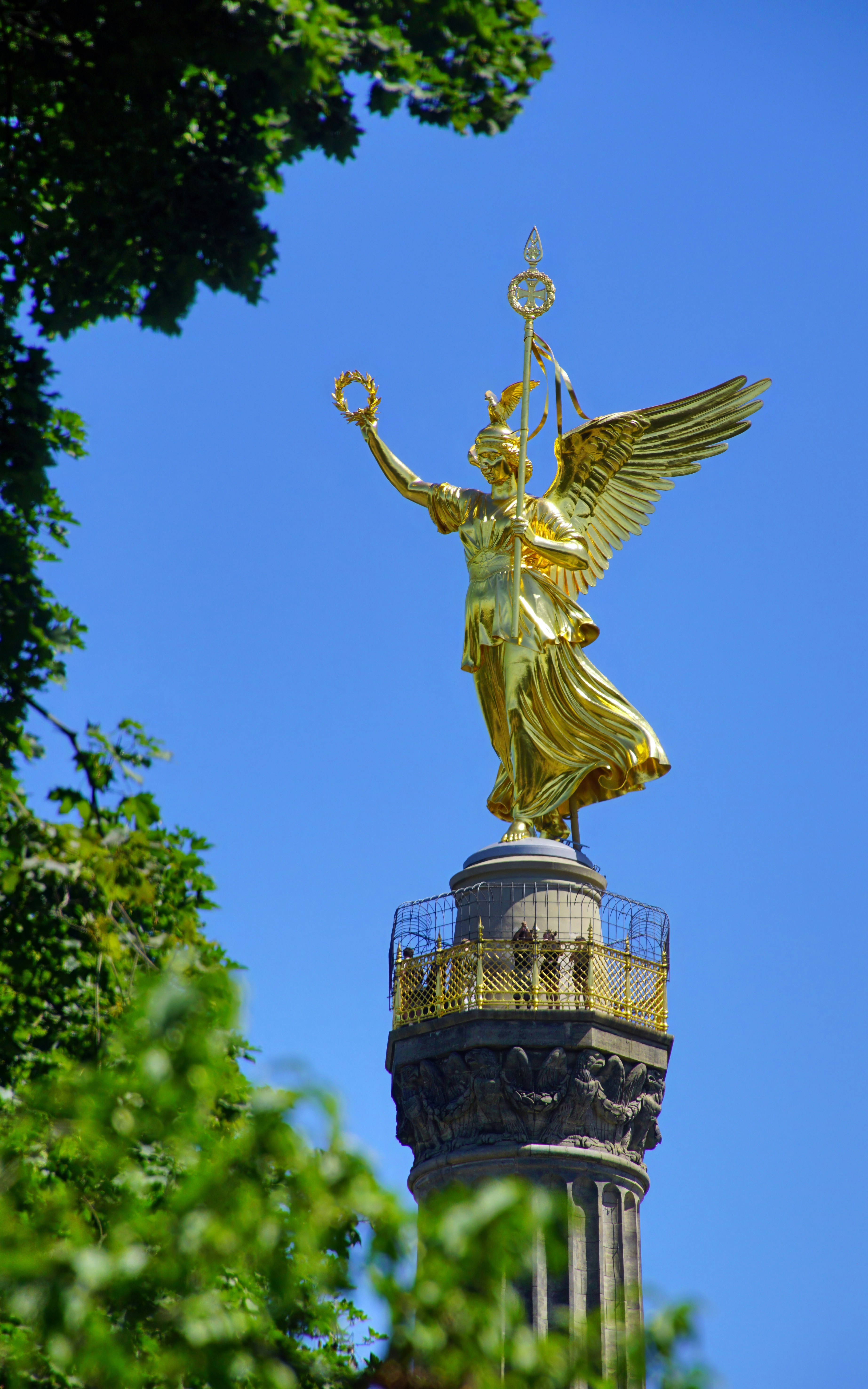 The Victory Column in Berlin on a sunny summer day. Designed by Heinrich Strack after 1864 to commemorate the Prussian victory over Denmark in the Second Schleswig War, by the time it was inaugurated in 1873, Prussia had also defeated Austria and its German allies in the Austro-Prussian War (1866) and France in the Franco-Prussian War (1870–71), giving the statue a new purpose. Different from the original plans, these later victories in the unification wars inspired the addition of the bronze sculpture of Victoria, the Roman goddess of victory, 8.3 metres (27 ft) high. | Golden angel statue atop column against blue sky.