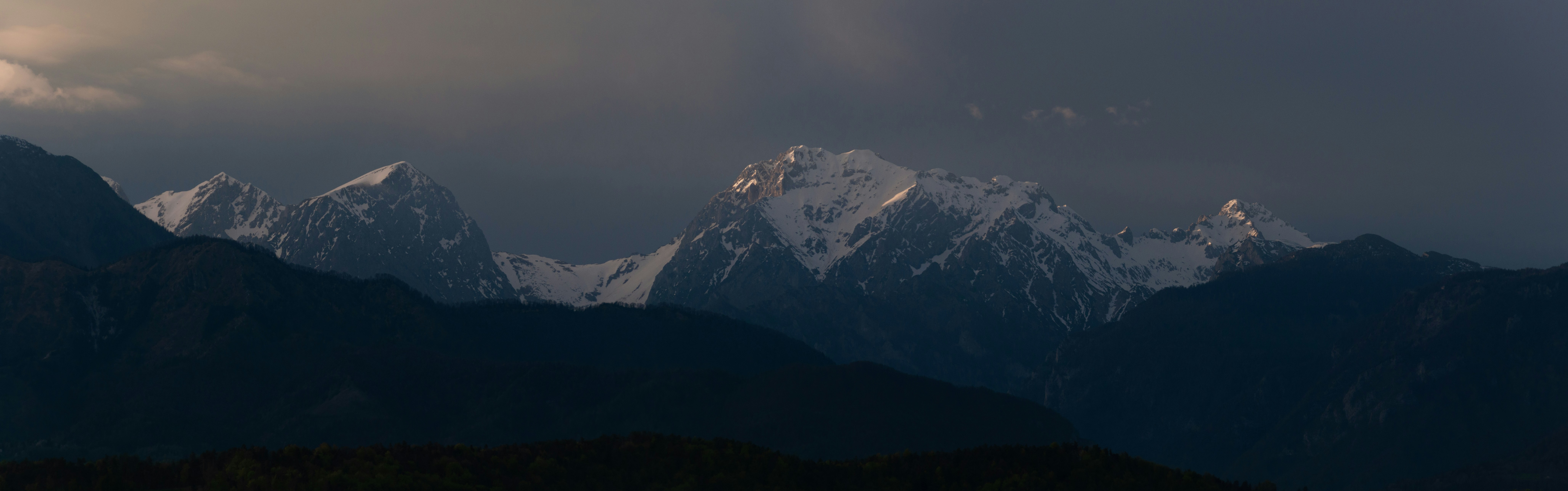 Majestic snow-capped mountains loom under a moody sky, their rugged silhouettes contrasting against the twilight. The scene captures the serene yet powerful essence of nature.