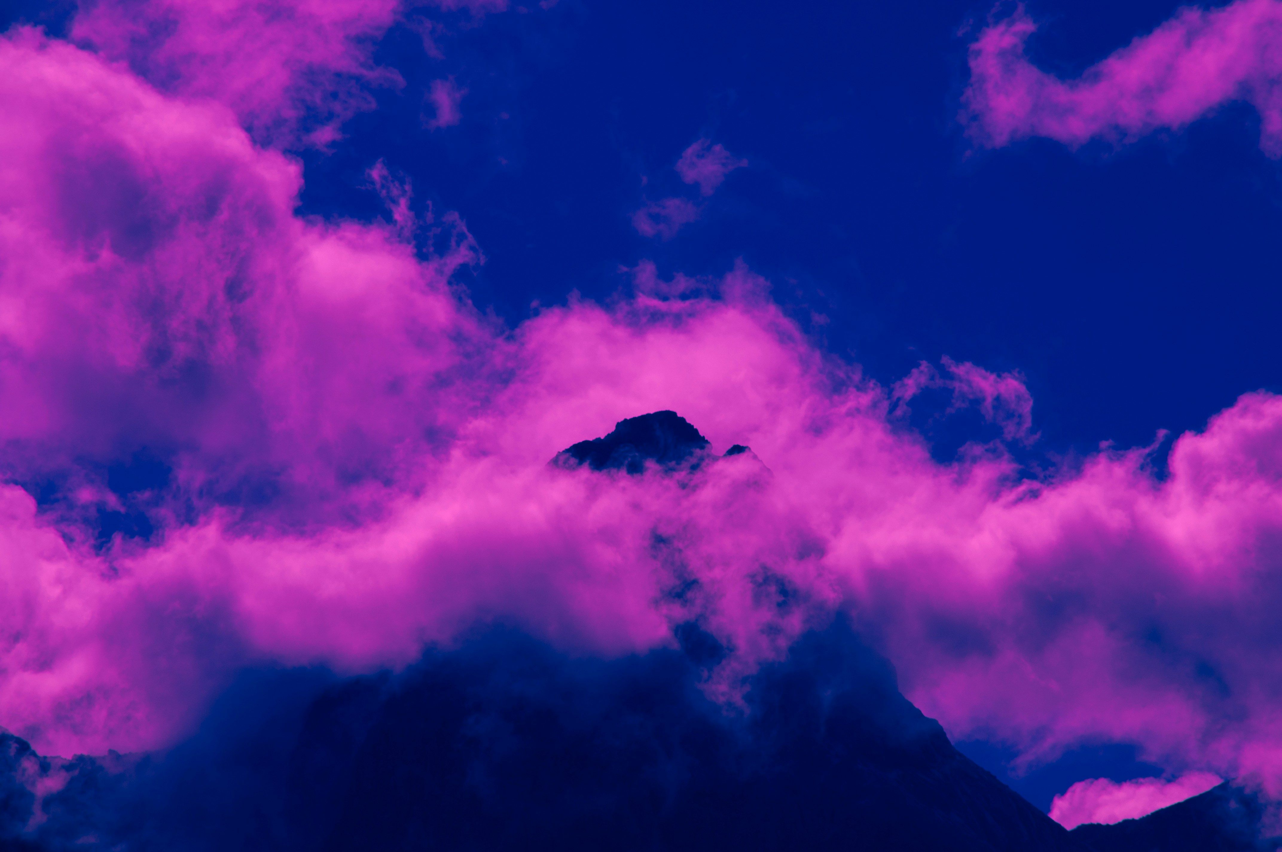 A striking, surreal landscape where the dark silhouette of a rugged mountain peak is dramatically framed by intensely vibrant magenta and pink clouds against a dark blue sky. A moody and powerful image of natural drama. | Dark mountain peak emerges through vibrant pink clouds.