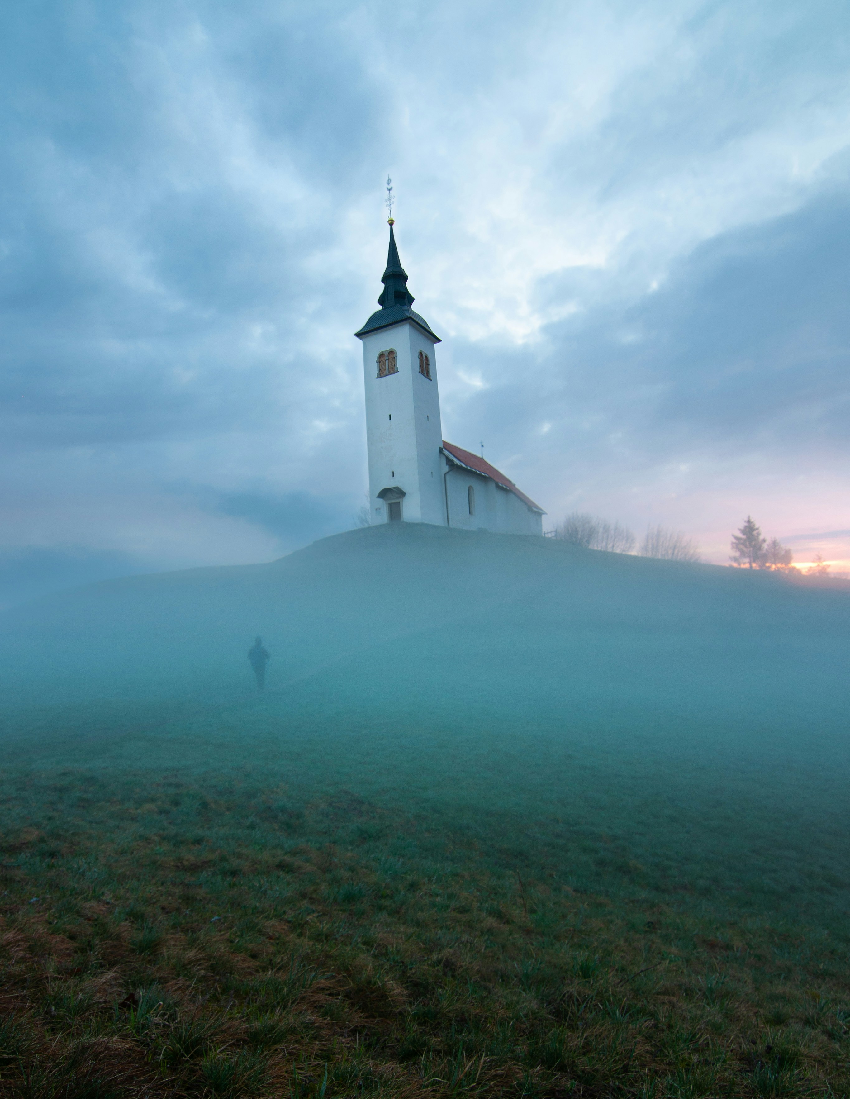 A dramatic, misty morning view of a white church with a tall steeple situated atop a grassy hill. The scene is enveloped in cool fog, creating a mystical and serene atmosphere as a solitary figure walks in the foreground. | White church on a hill shrouded in fog.