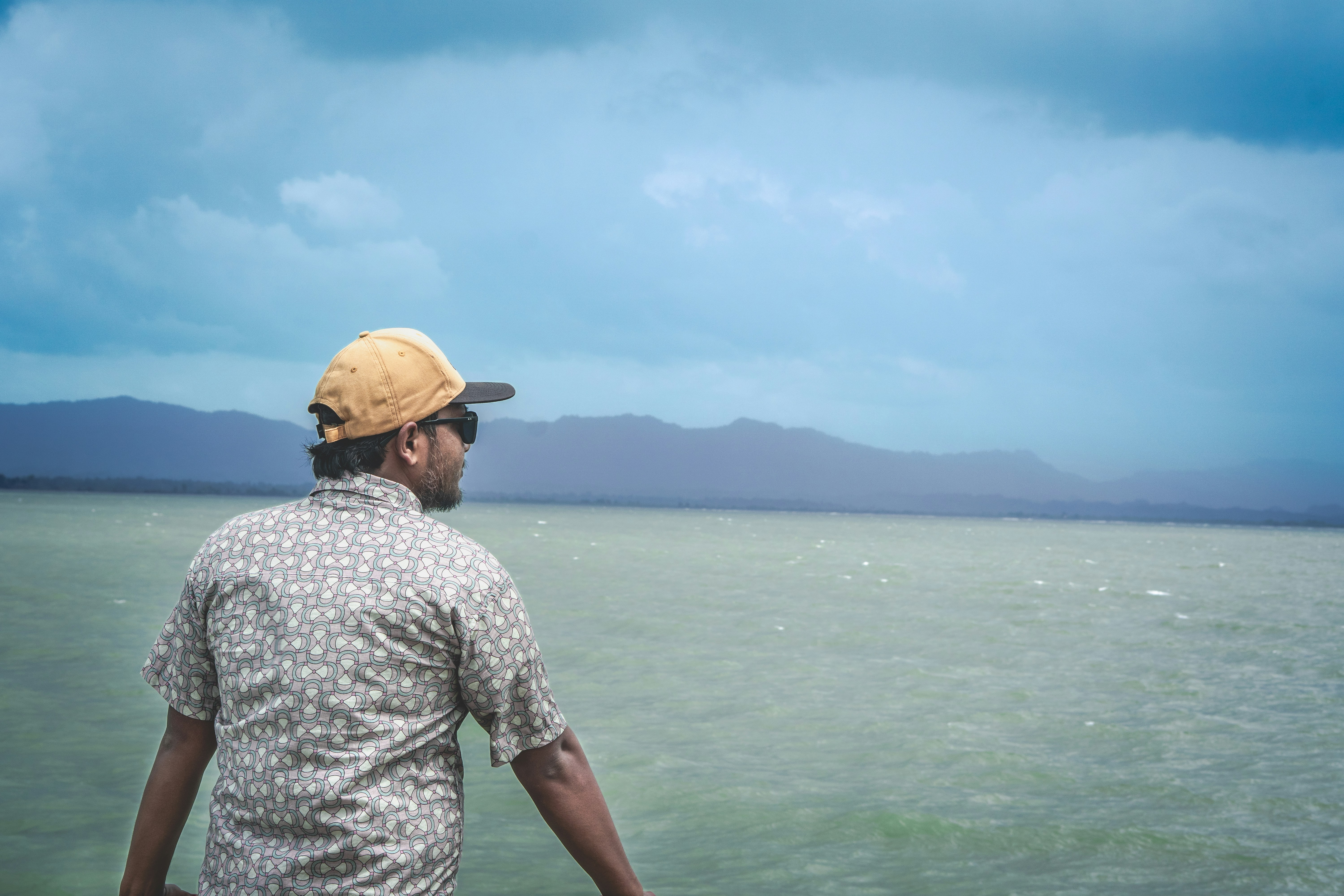 A man stands by a vast, calm sea, gazing toward the distant mountains under a dramatic blue sky. He wears a patterned shirt, a tan cap, and sunglasses, creating a relaxed yet adventurous vibe. The scene beautifully captures the essence of solitude, reflection, and wanderlust — the calm water meeting the cloudy horizon evokes a feeling of peace and contemplation. The natural hues of the sea and sky make this image perfect for travel, lifestyle, and nature photography themes. | Man in hat looks at distant mountains over water.