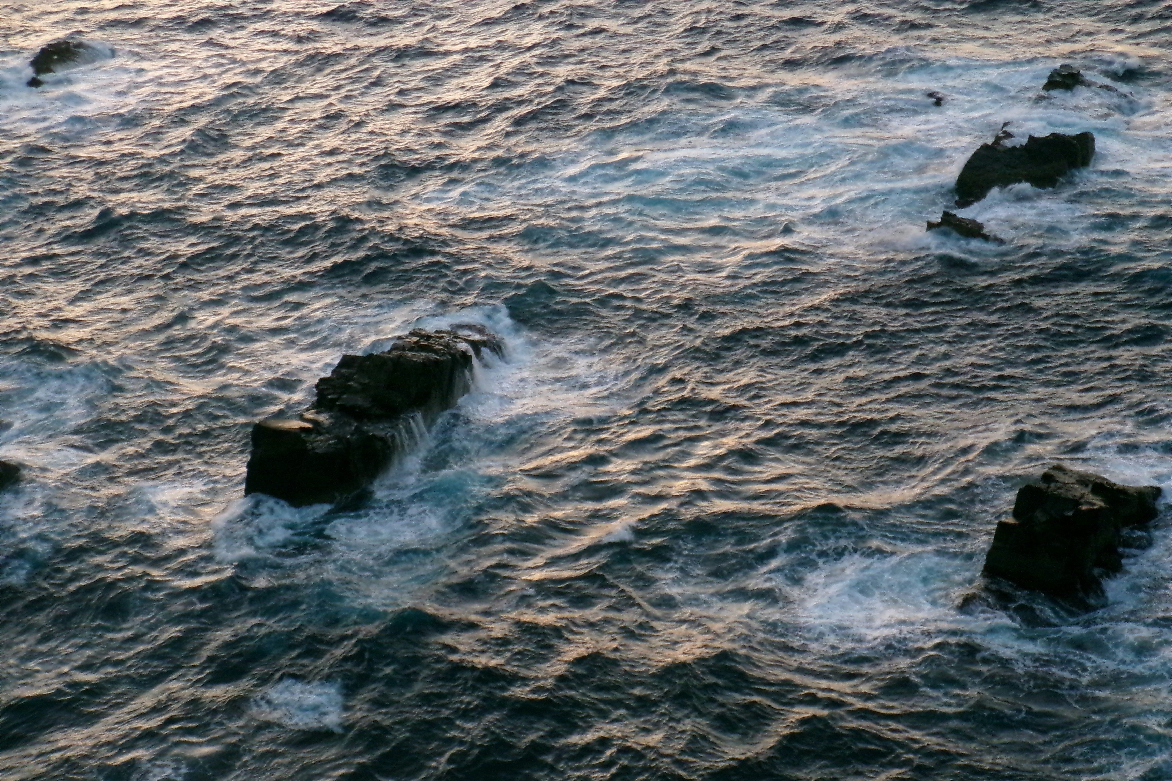 Rugged rocks emerge from the gently swirling ocean, kissed by the soft hues of twilight. The interplay of water and stone creates a serene coastal scene.