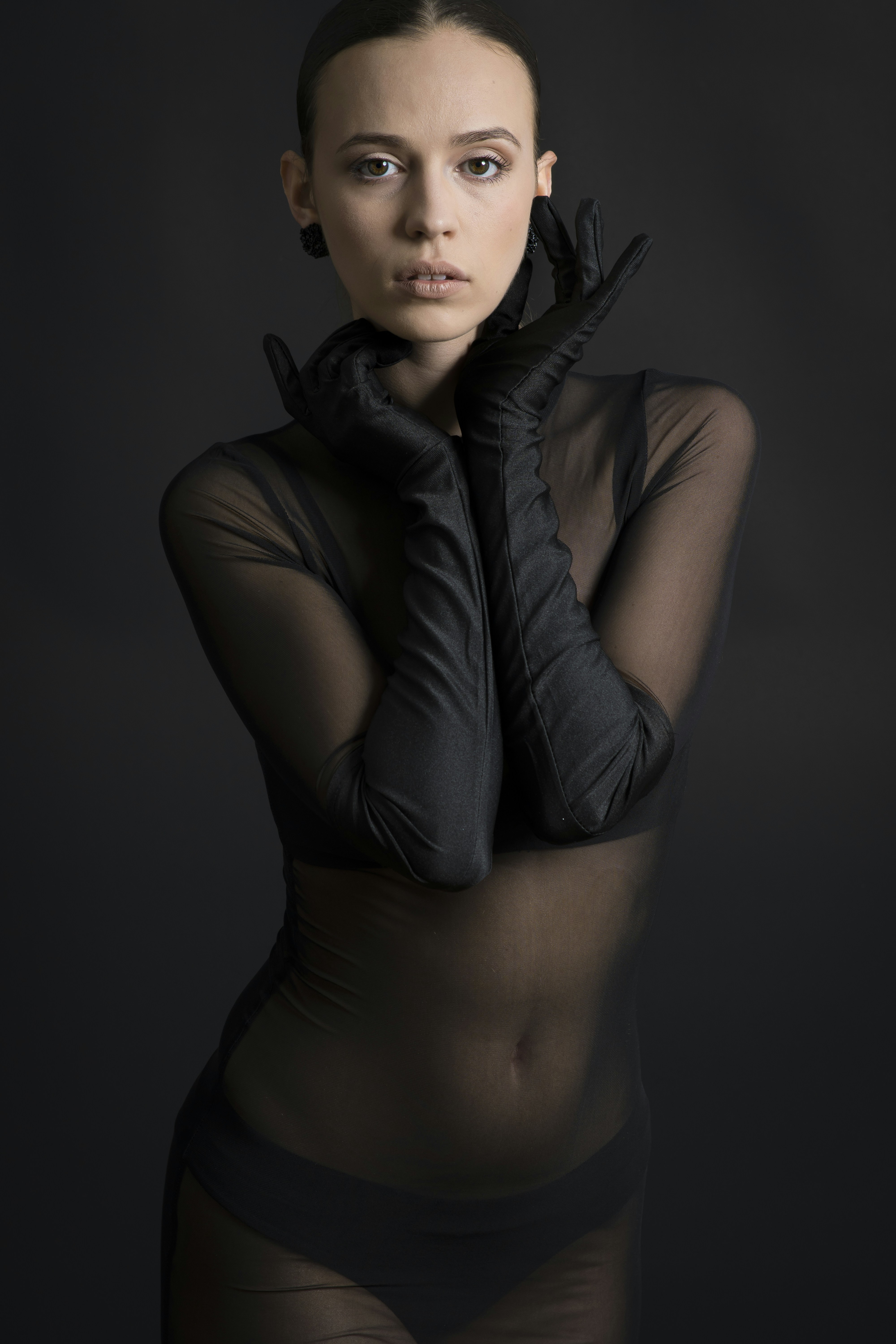 Model in sheer black attire striking a pose against a dark backdrop, emphasizing the interplay of light and shadow.