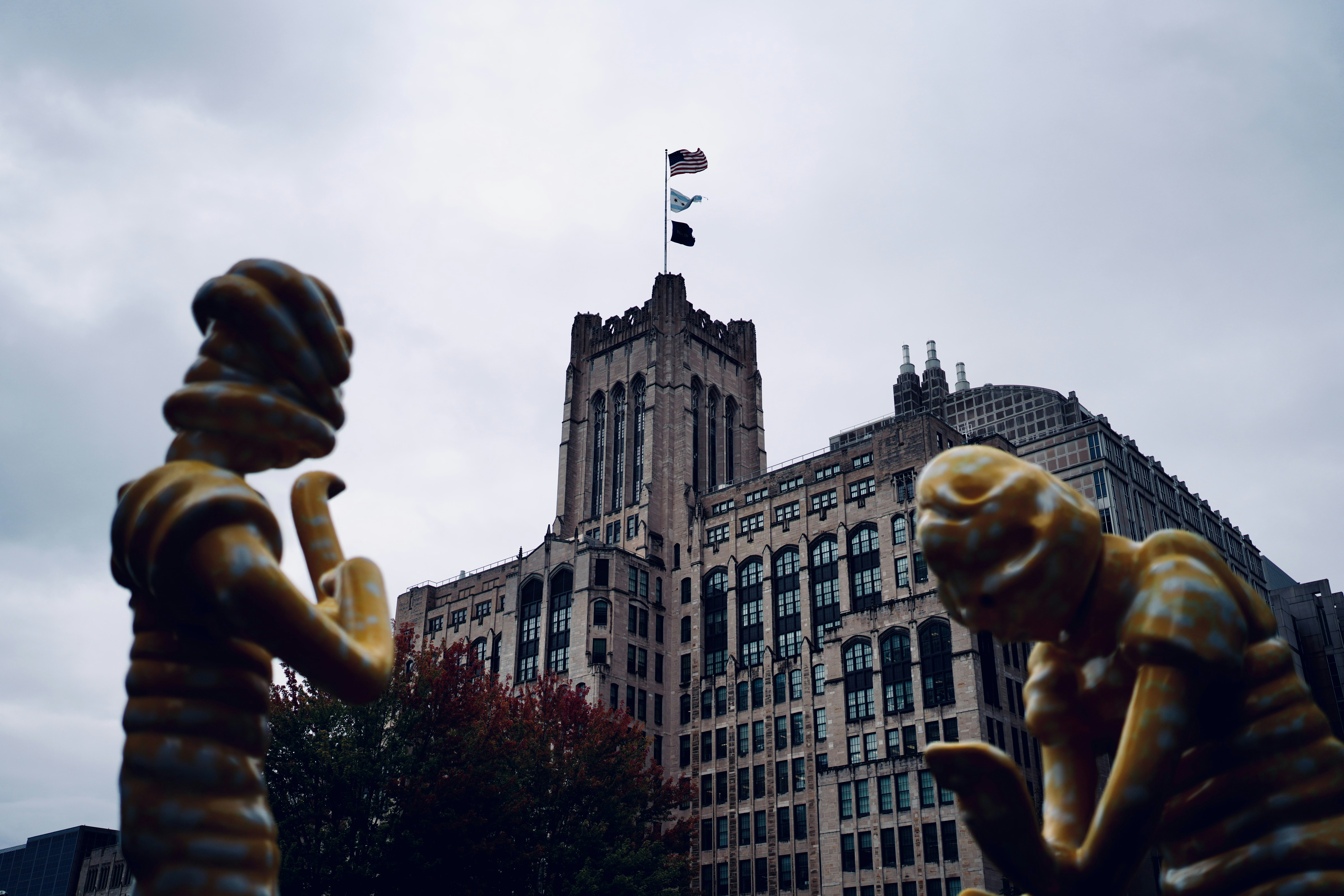 Two abstract yellow sculptures in the foreground, with a historic building and flags in the background under a cloudy sky.