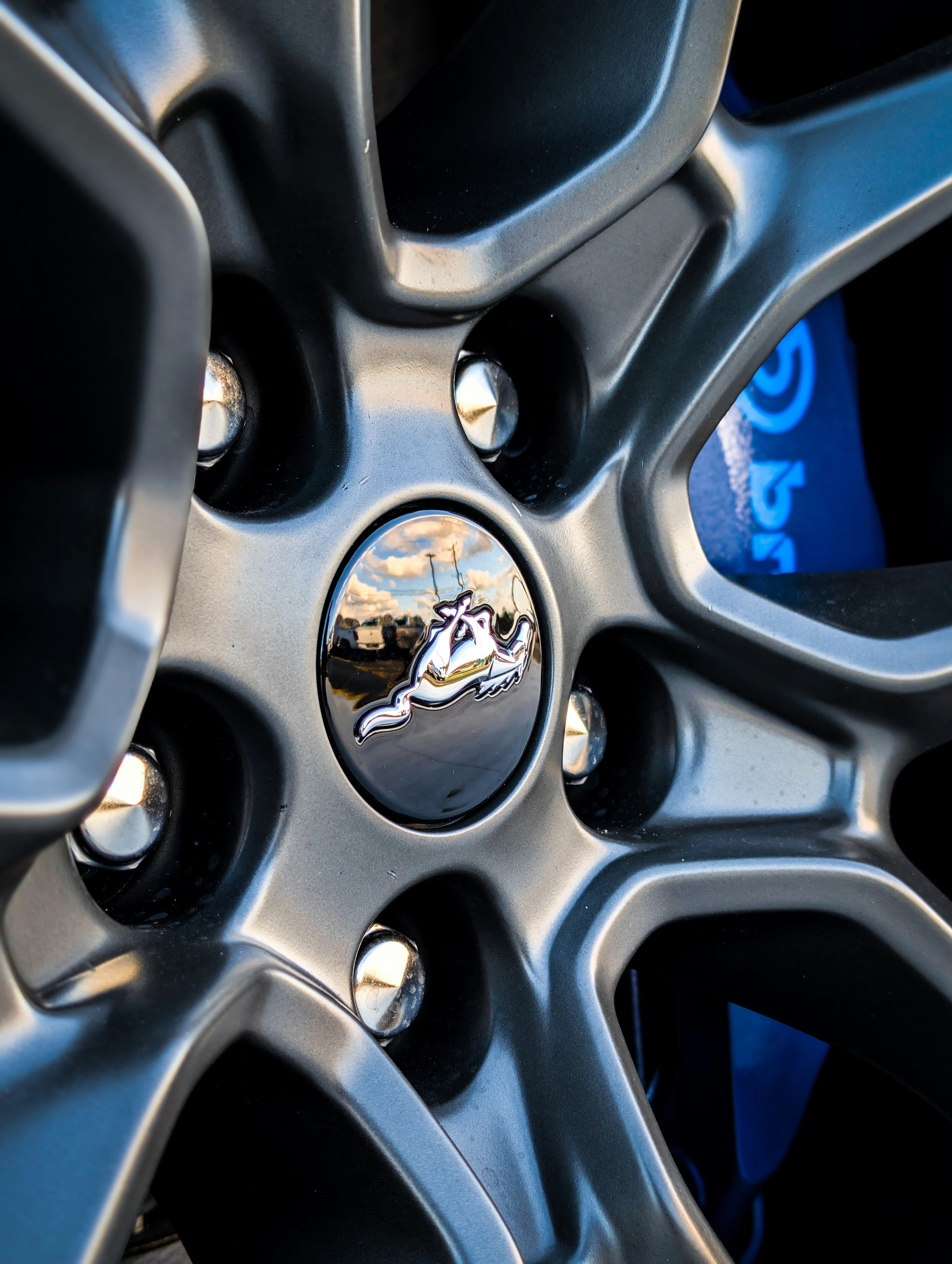 A stunning macro view where the iconic Running Pony emblem, gleaming with reflections, is beautifully framed by the matte graphite spokes. This close-up perfectly highlights the powerful synthesis of style and high-performance engineering. | Close-up of a car wheel with a horse emblem.
