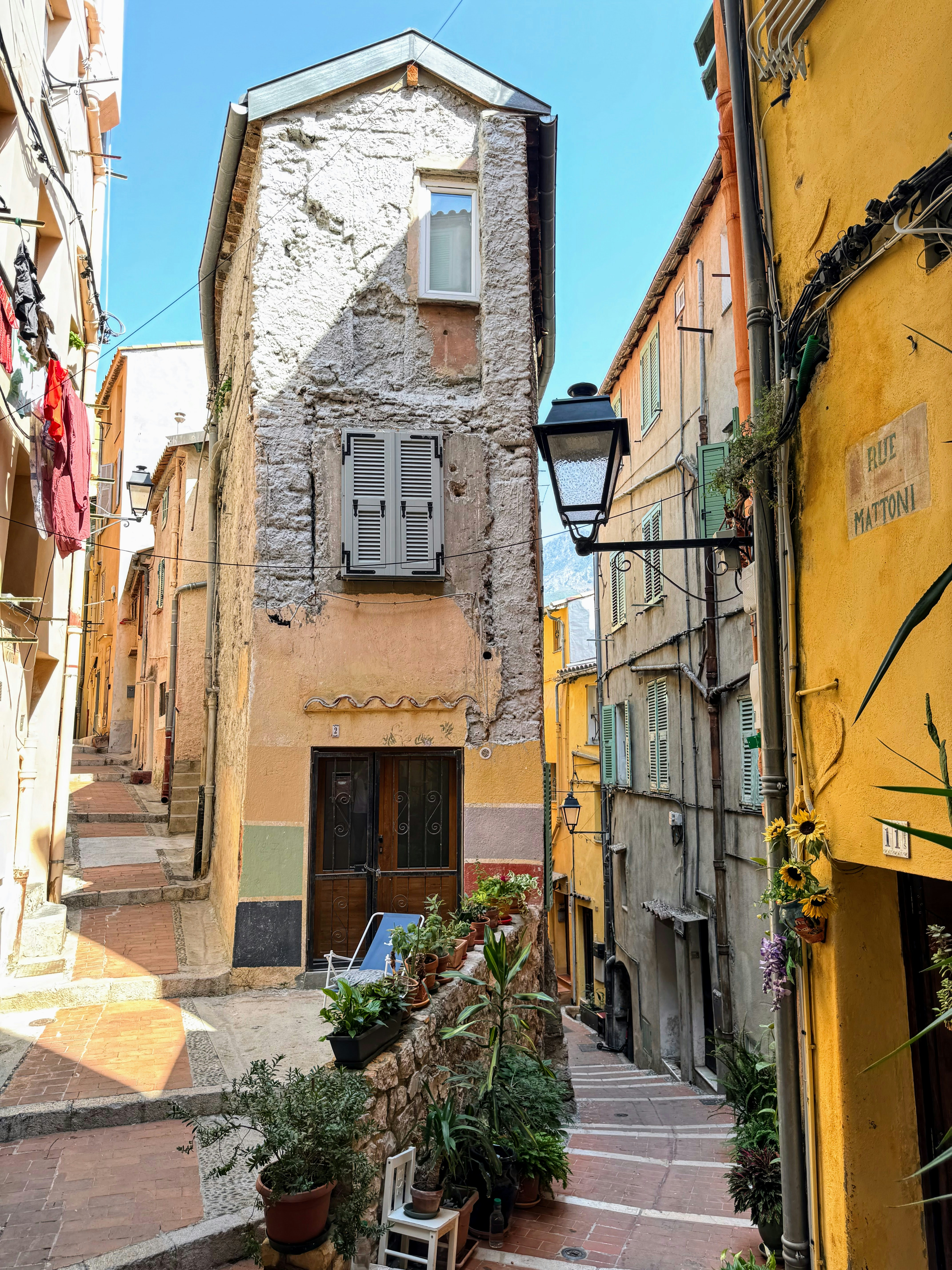 Narrow alleyway adorned with vibrant yellow walls and a charming stone building, featuring potted plants and hanging laundry. A vintage street lamp adds to the quaint atmosphere.