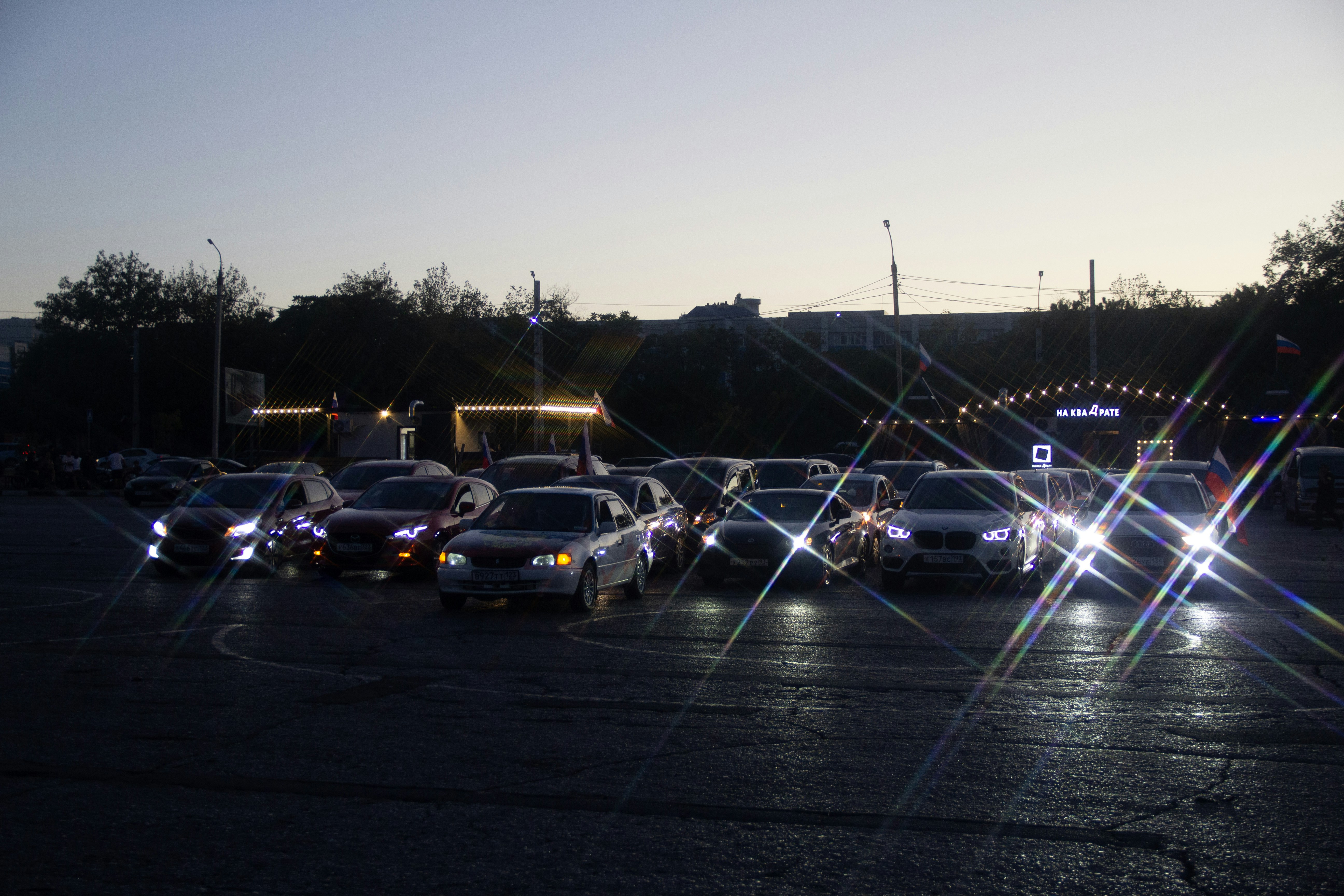 Cars lined up in a parking lot at dusk, their headlights creating a sparkling effect against the darkening sky.