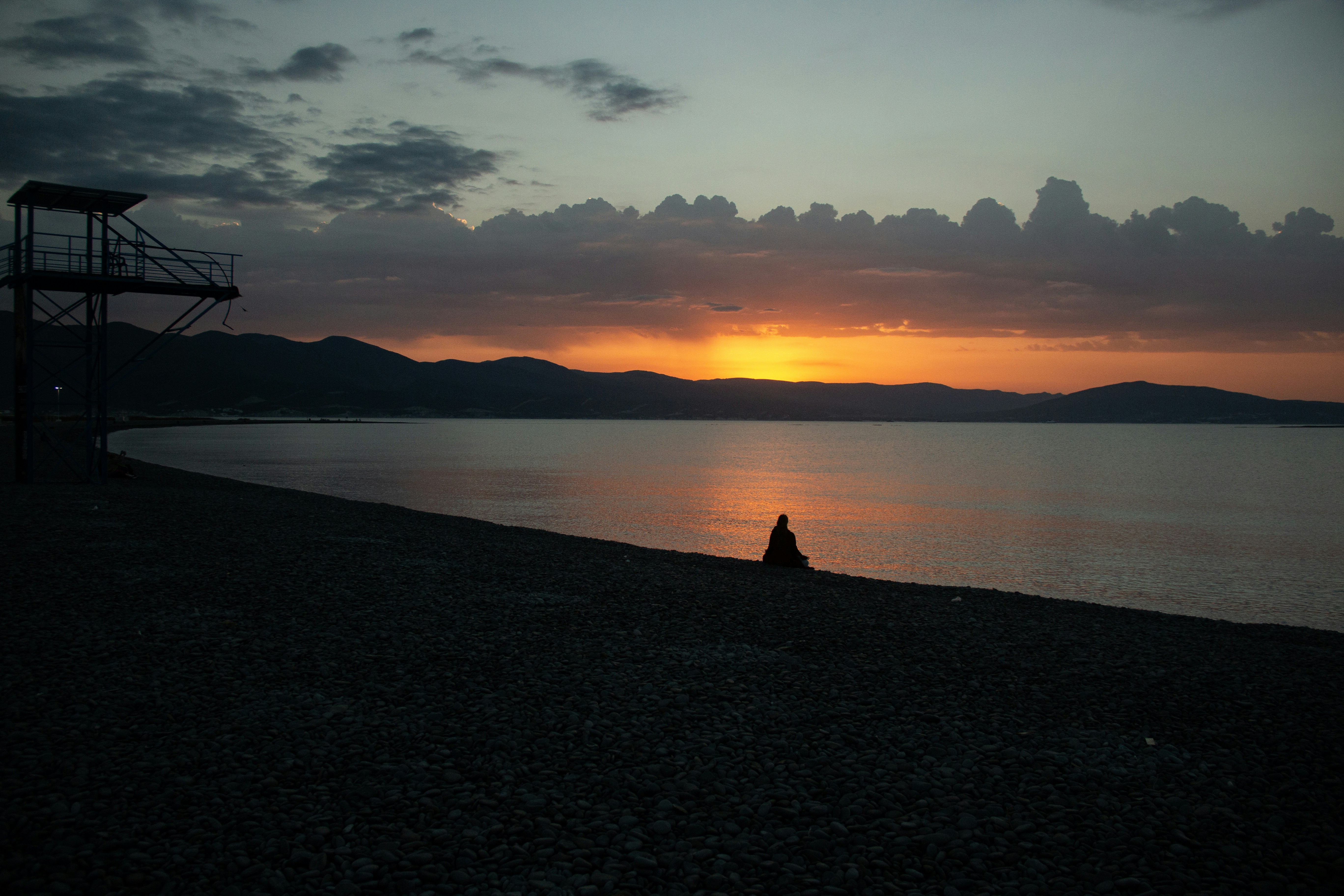Silhouetted figure seated on a rocky beach, gazing at the vibrant sunset over calm waters with distant mountains. 