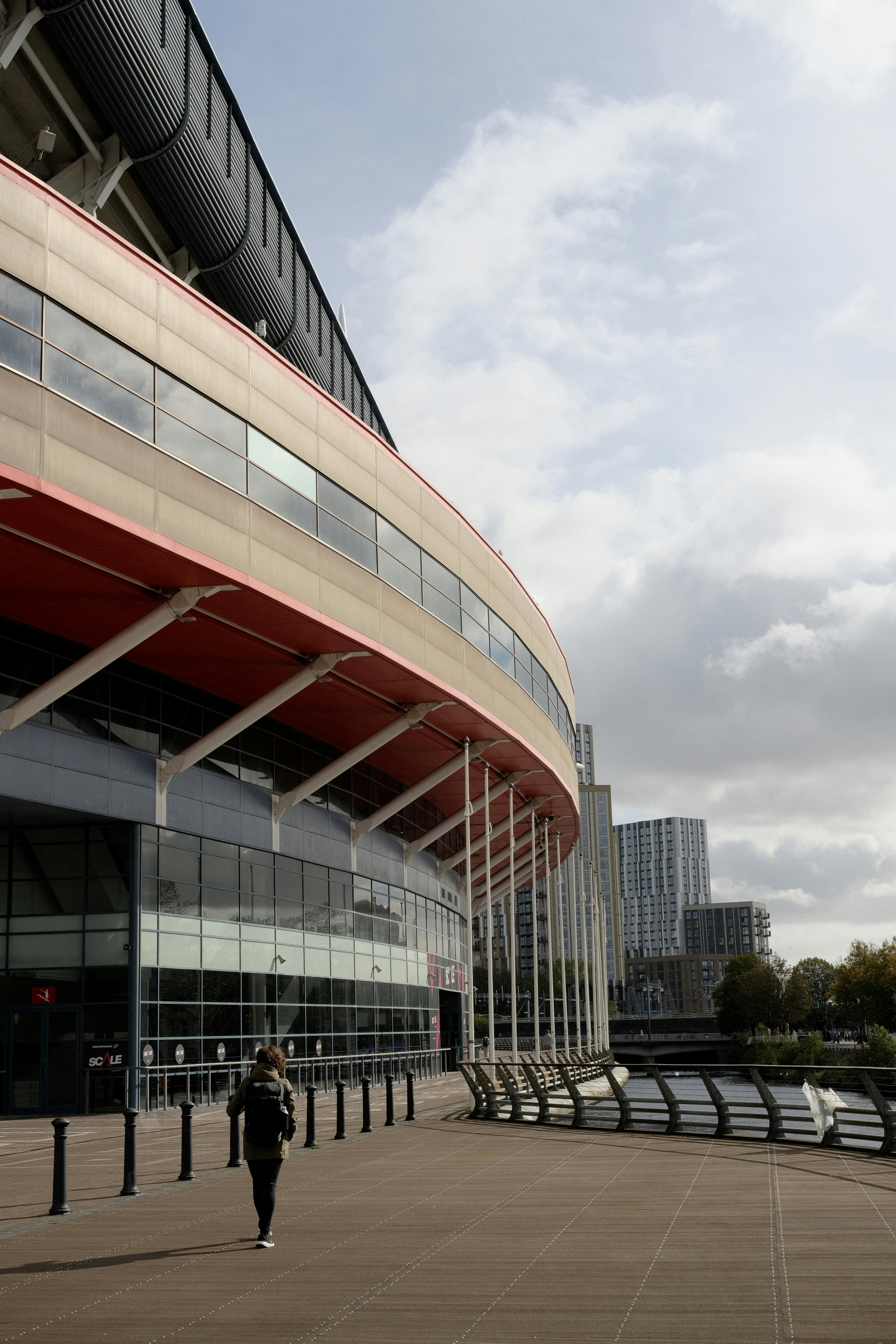 A lone figure walks along a riverside pathway, framed by the sweeping curves of a contemporary building and the skyline of a bustling city.