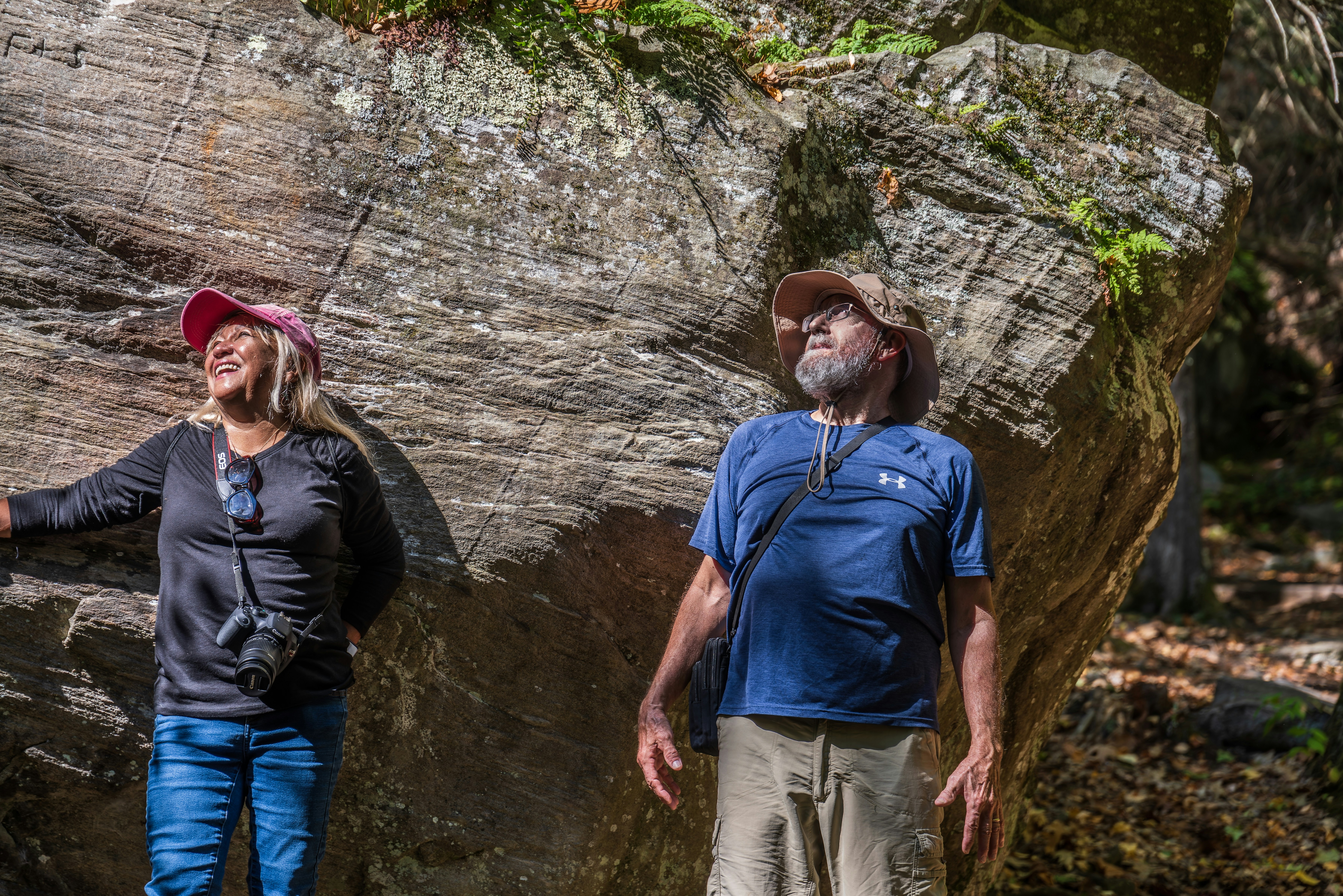 Two hikers express joy and wonder beneath a towering rock formation in a sunlit forest. The scene captures the essence of outdoor adventure.