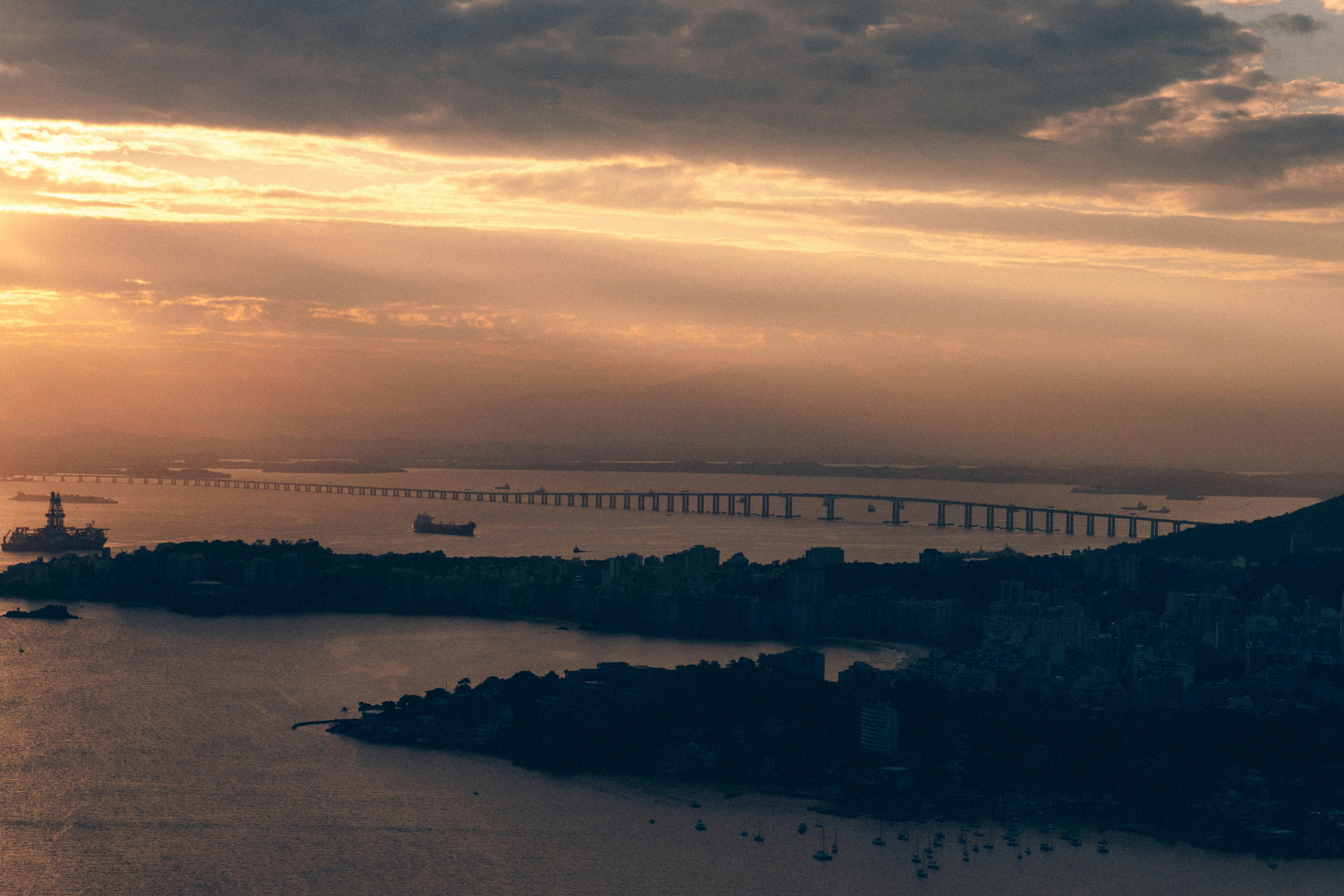 A tranquil view of a bridge stretching across the water at sunset, with silhouettes of buildings and boats in the foreground. The soft glow of the sky adds depth to the scene.