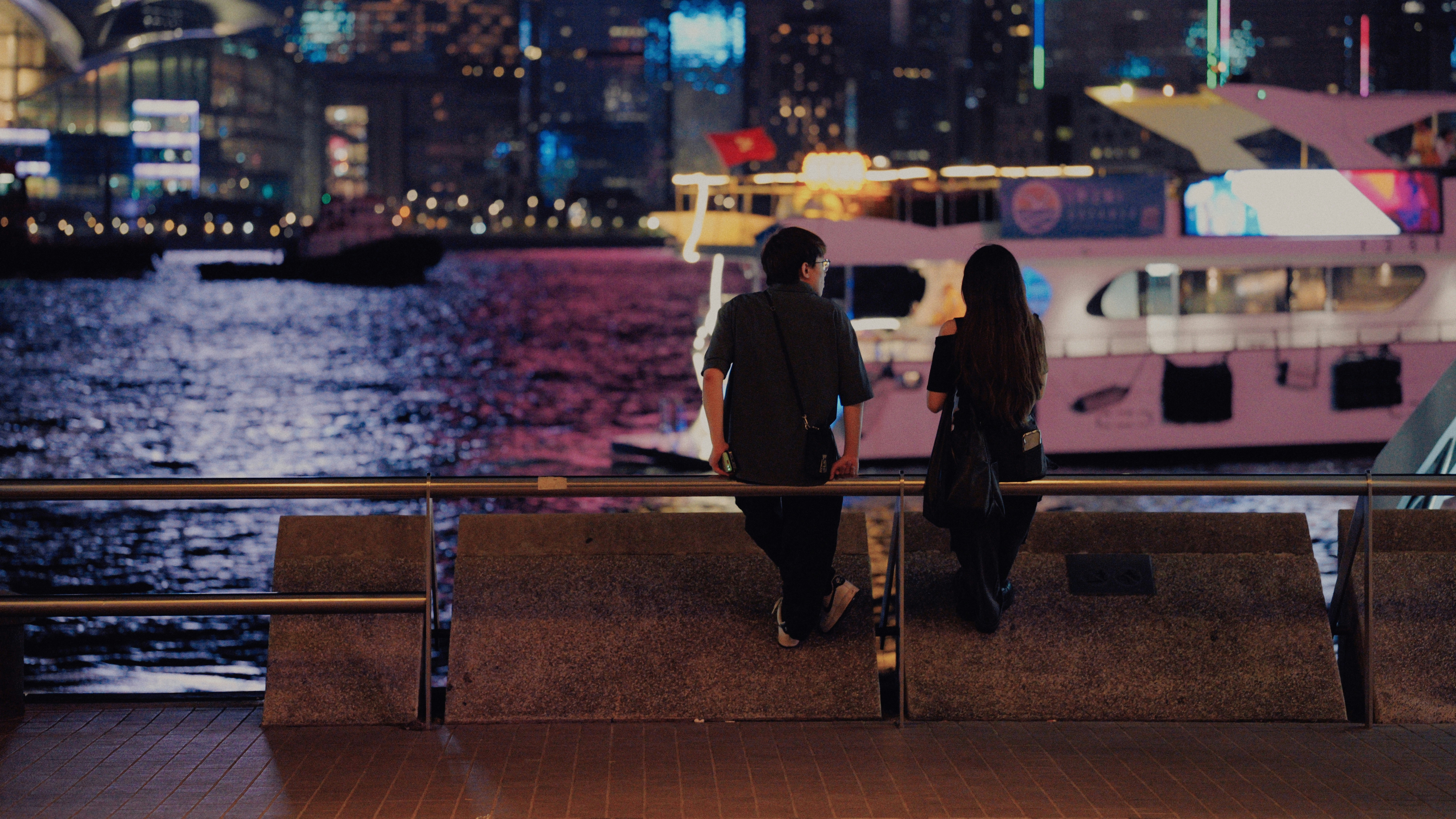 Two figures seated on a waterfront barrier, gazing at illuminated boats and city lights reflecting on the water.