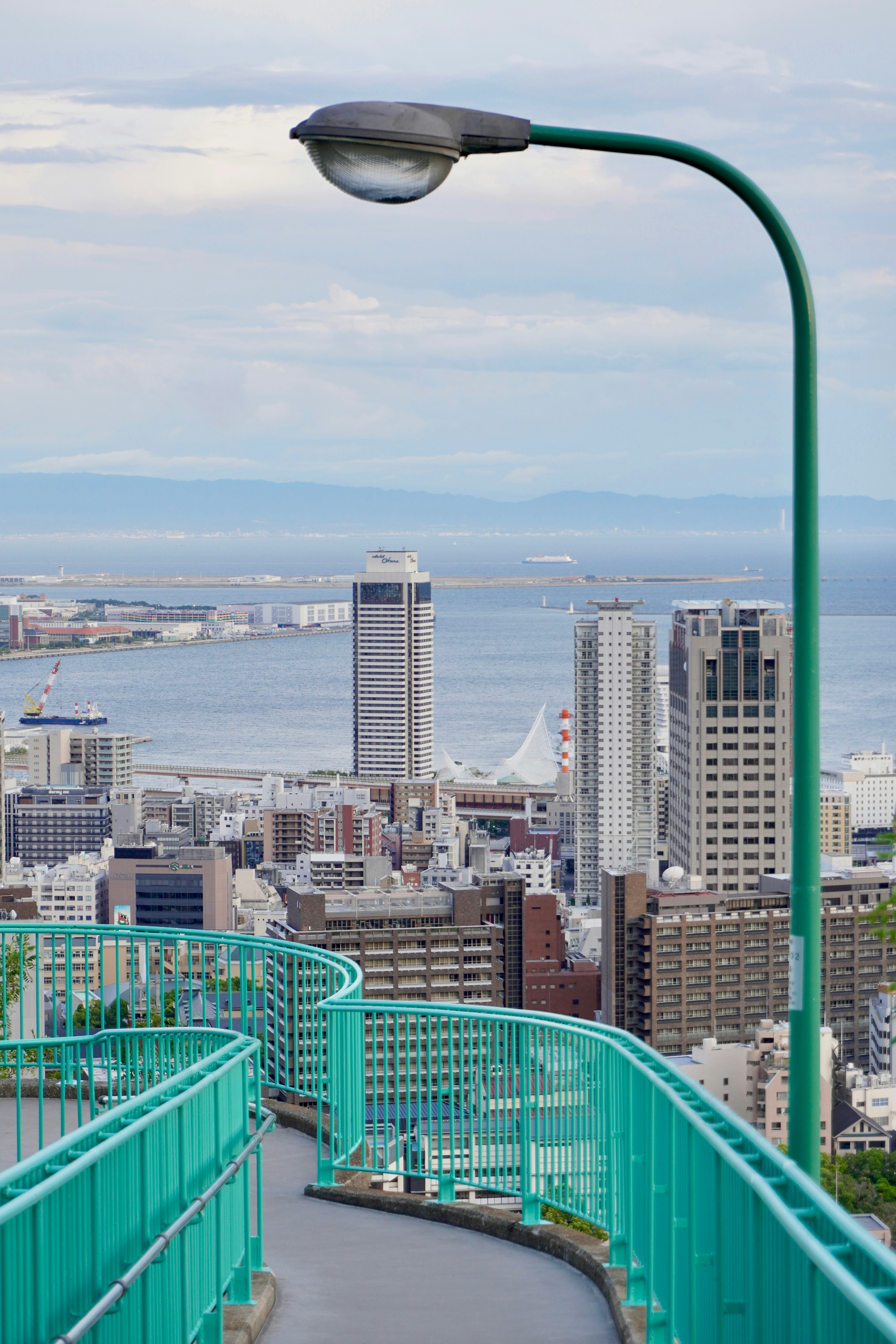 A winding pathway leads to a panoramic view of a bustling coastal city, with modern skyscrapers and the shimmering sea in the background.