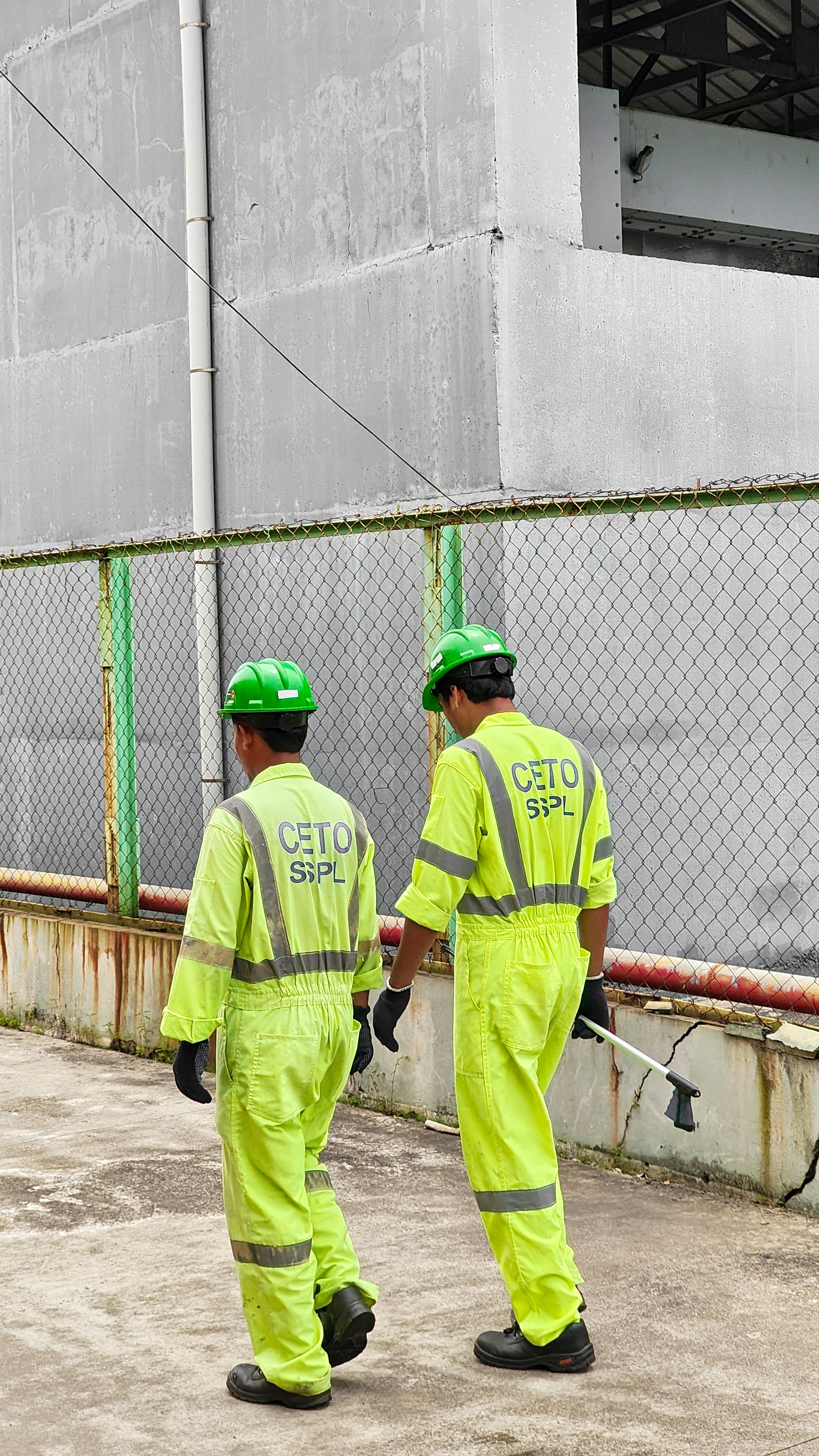 Two workers in neon safety suits walk past a fence.