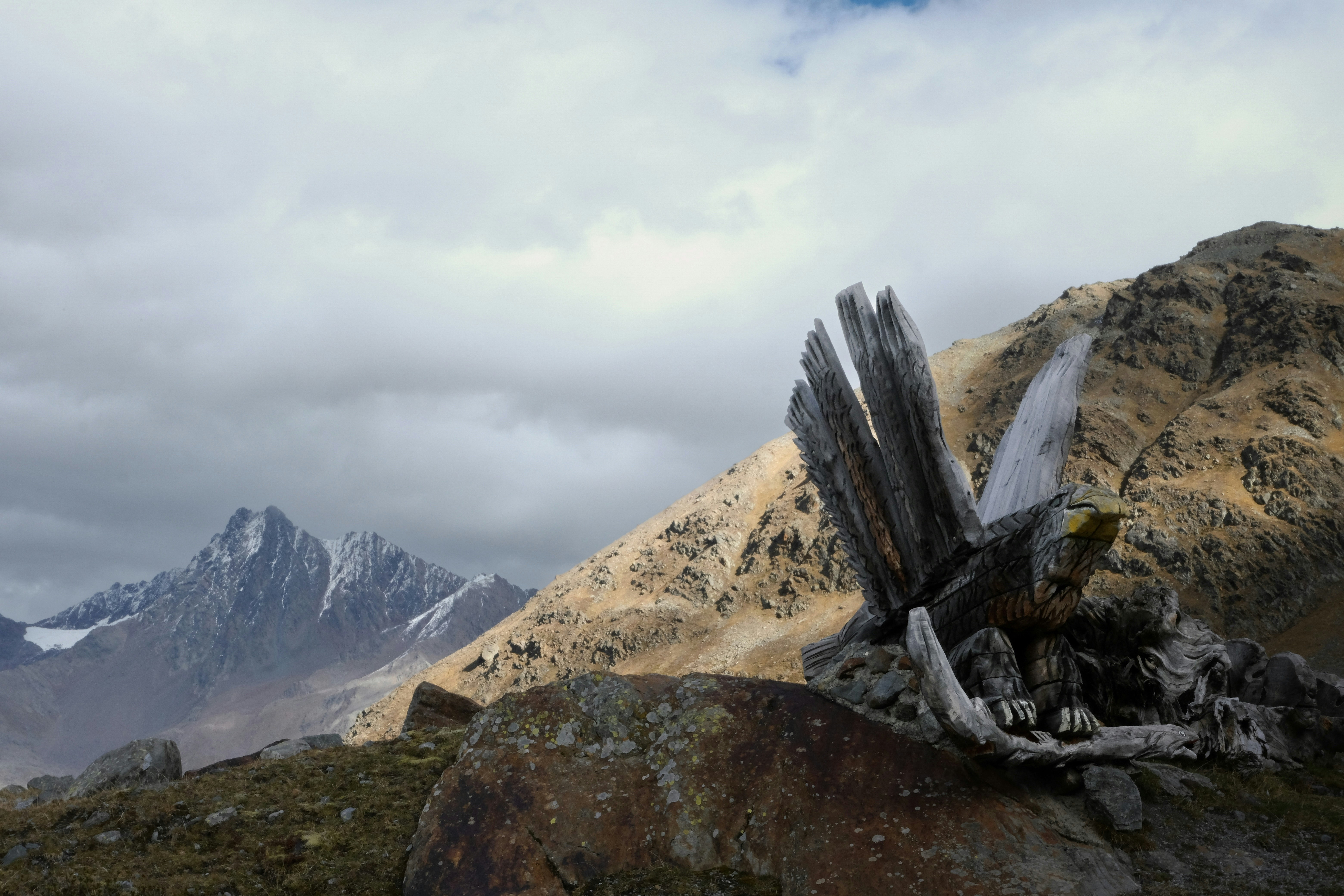 A majestic wooden sculpture resembling a mythical creature rests on a rocky outcrop, framed by towering mountains under a dynamic sky.