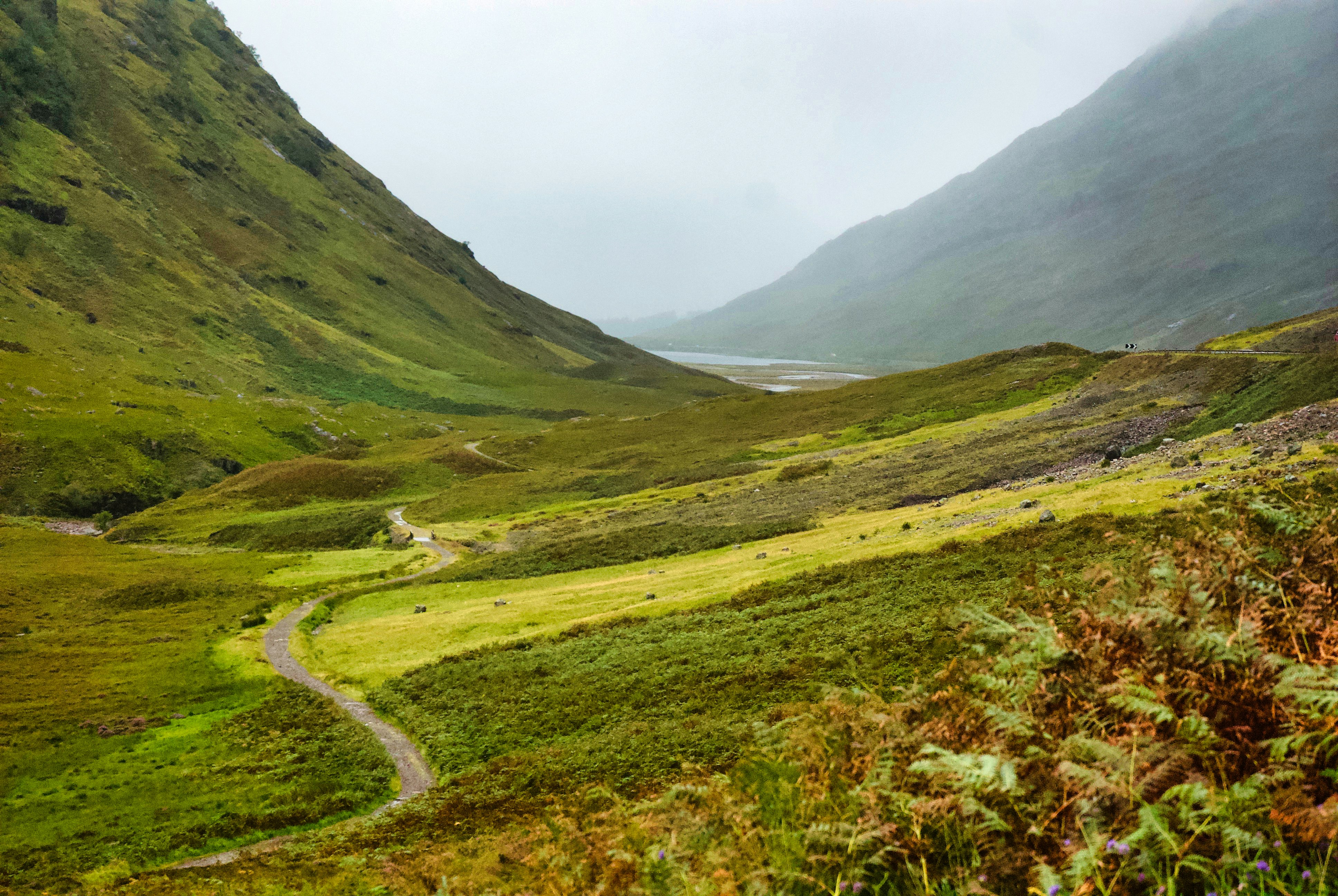 Lush green valley winding through the misty highlands, with a meandering path leading towards the distant waterway.