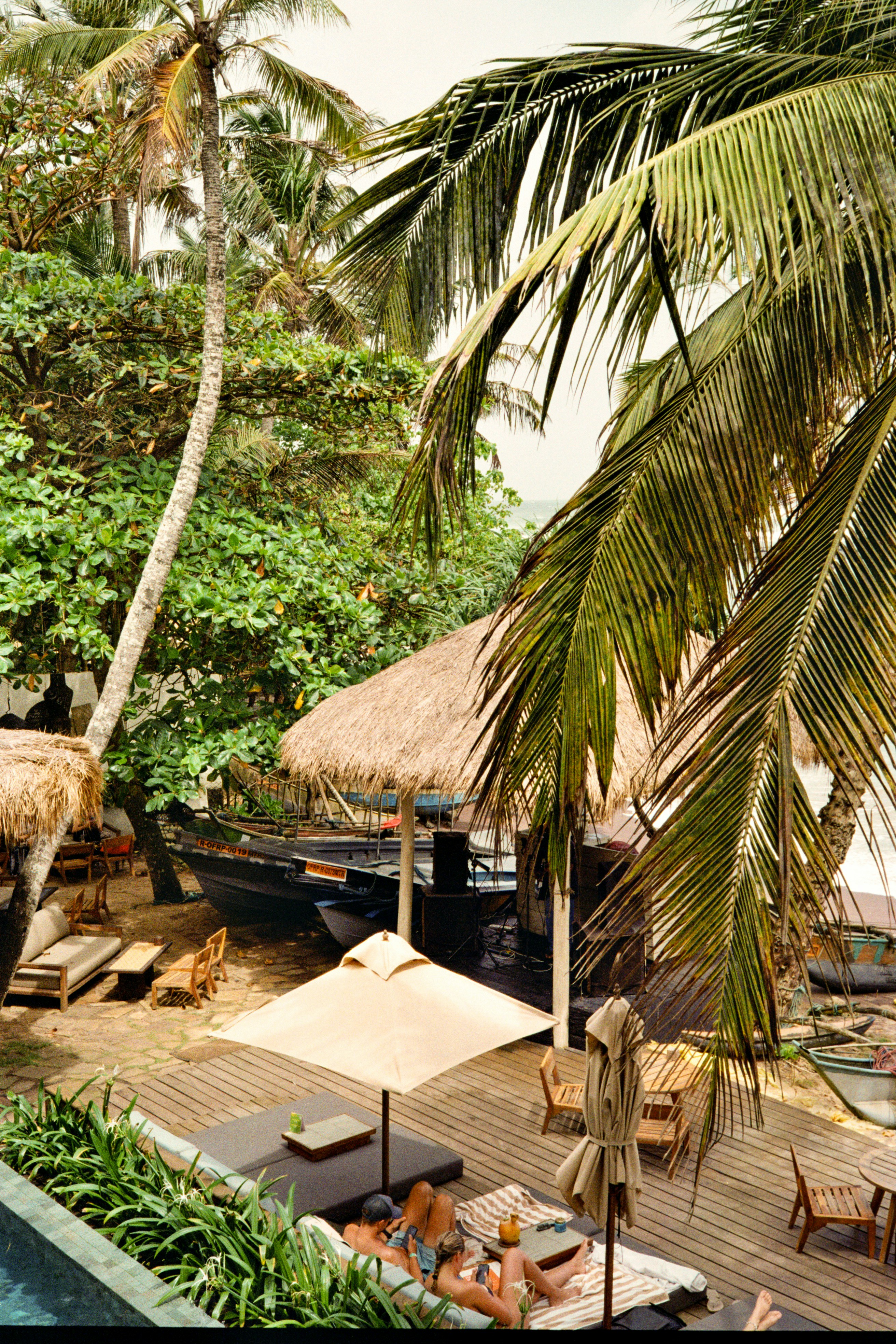 Lush greenery surrounds a cozy beachside retreat with sun loungers and a thatched roof, inviting relaxation. A sailboat is anchored nearby, adding to the tranquil atmosphere.